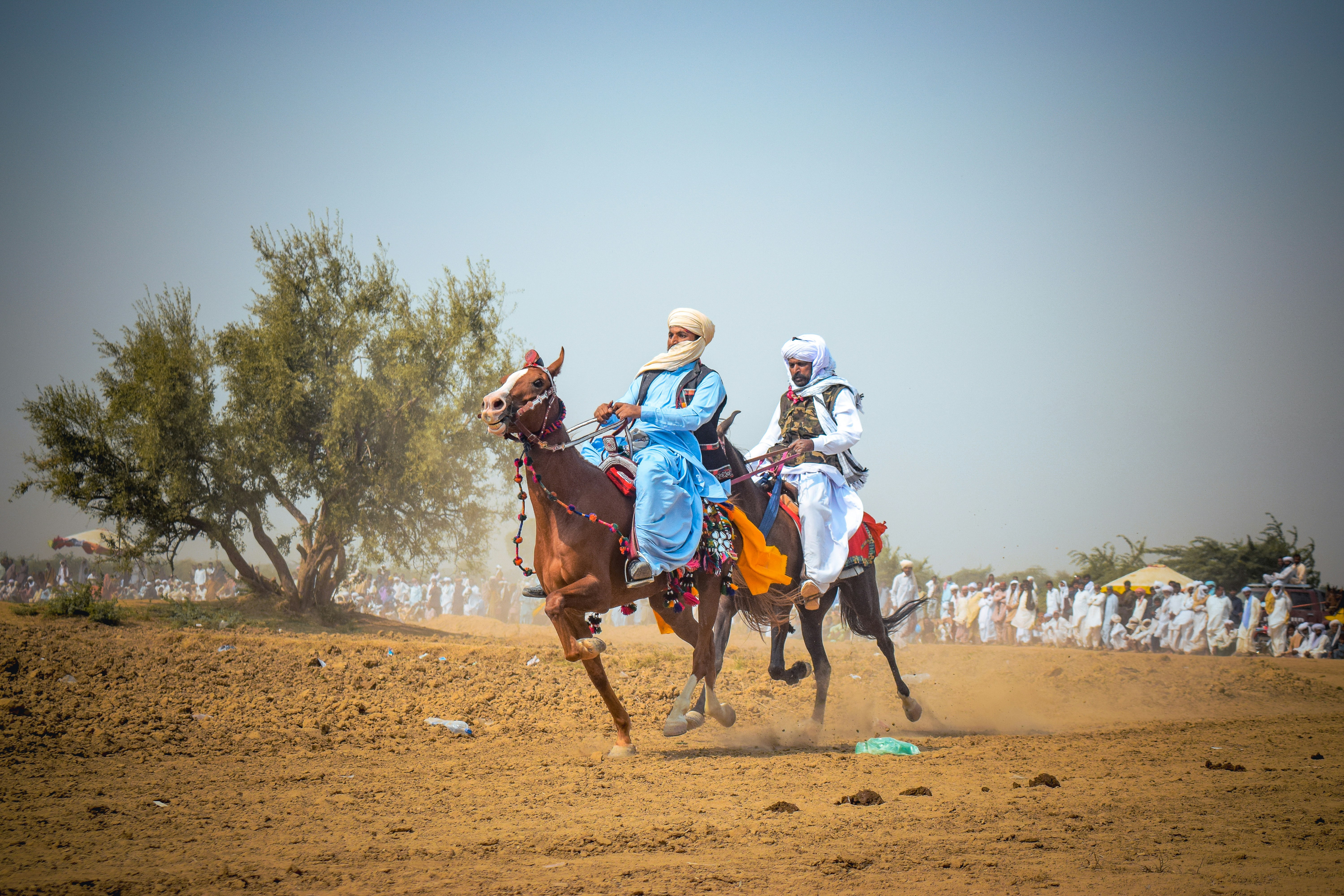 Two riders in traditional clothing race on horseback across a sandy landscape under a clear blue sky.
