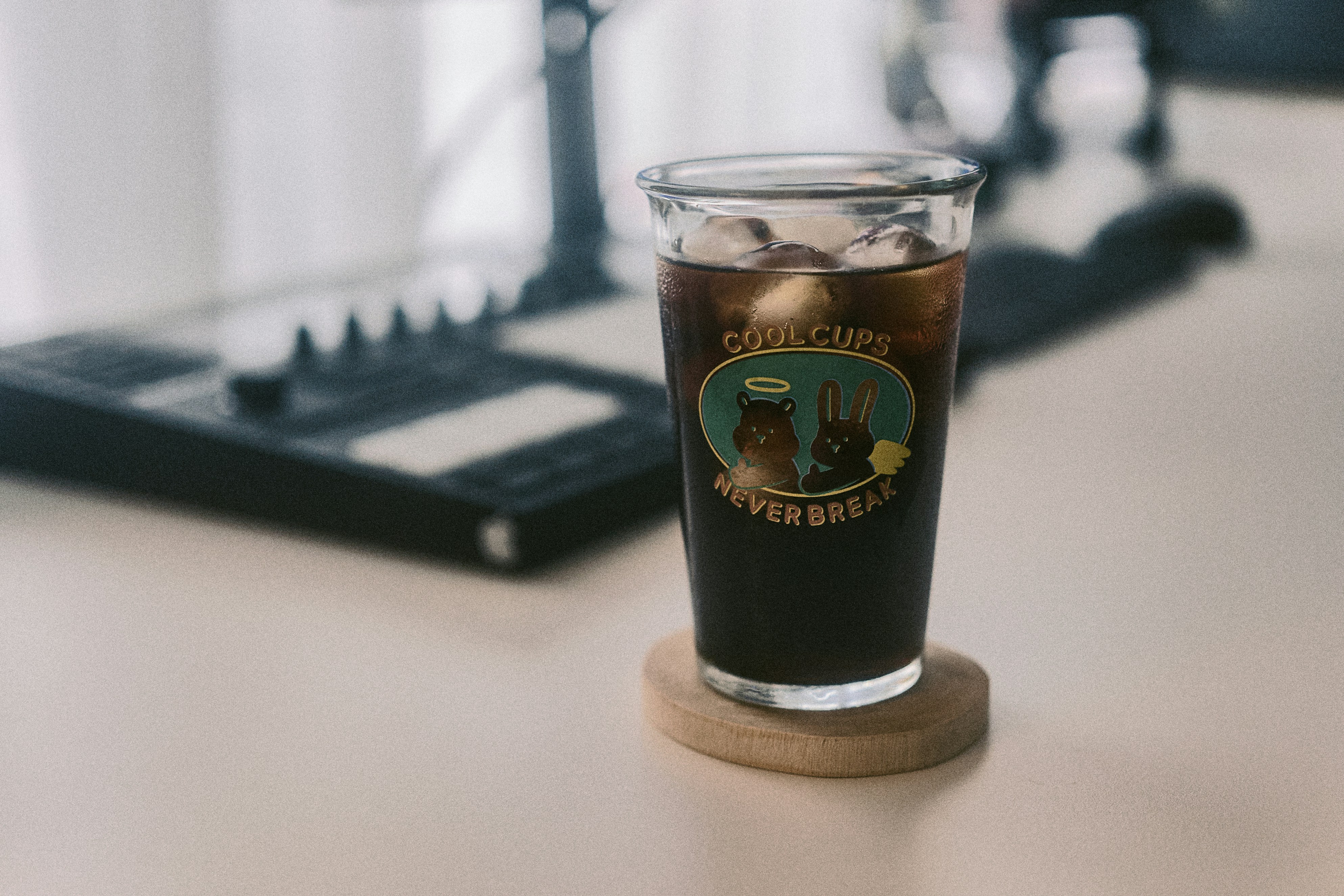 A photograph of a logo-bearing glass of dark cold brew on a wooden coaster, with a blurred keyboard in the background.