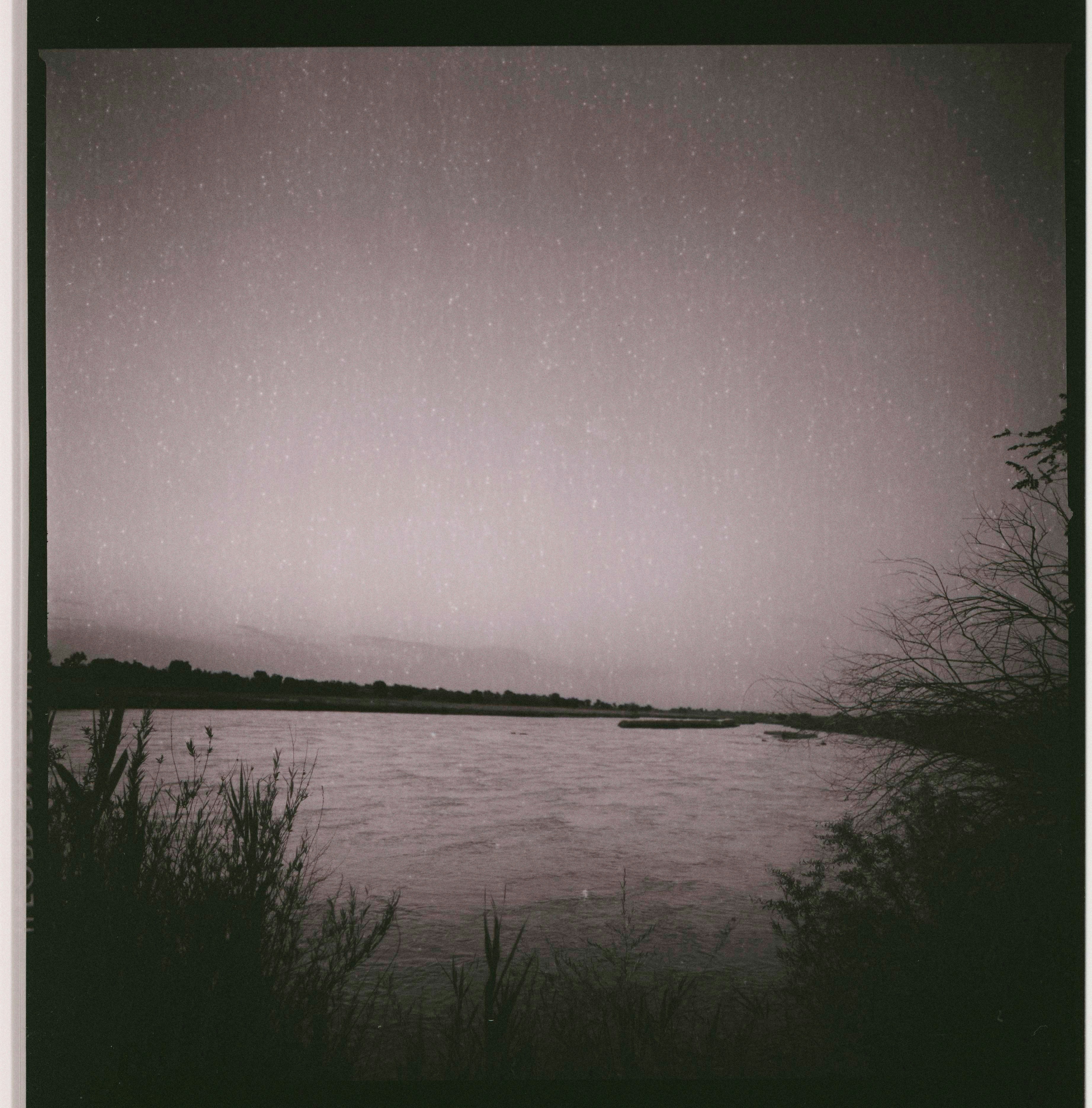 Serene riverbank under a twilight sky, with gentle ripples reflecting the fading light. Lush vegetation frames the scene.
