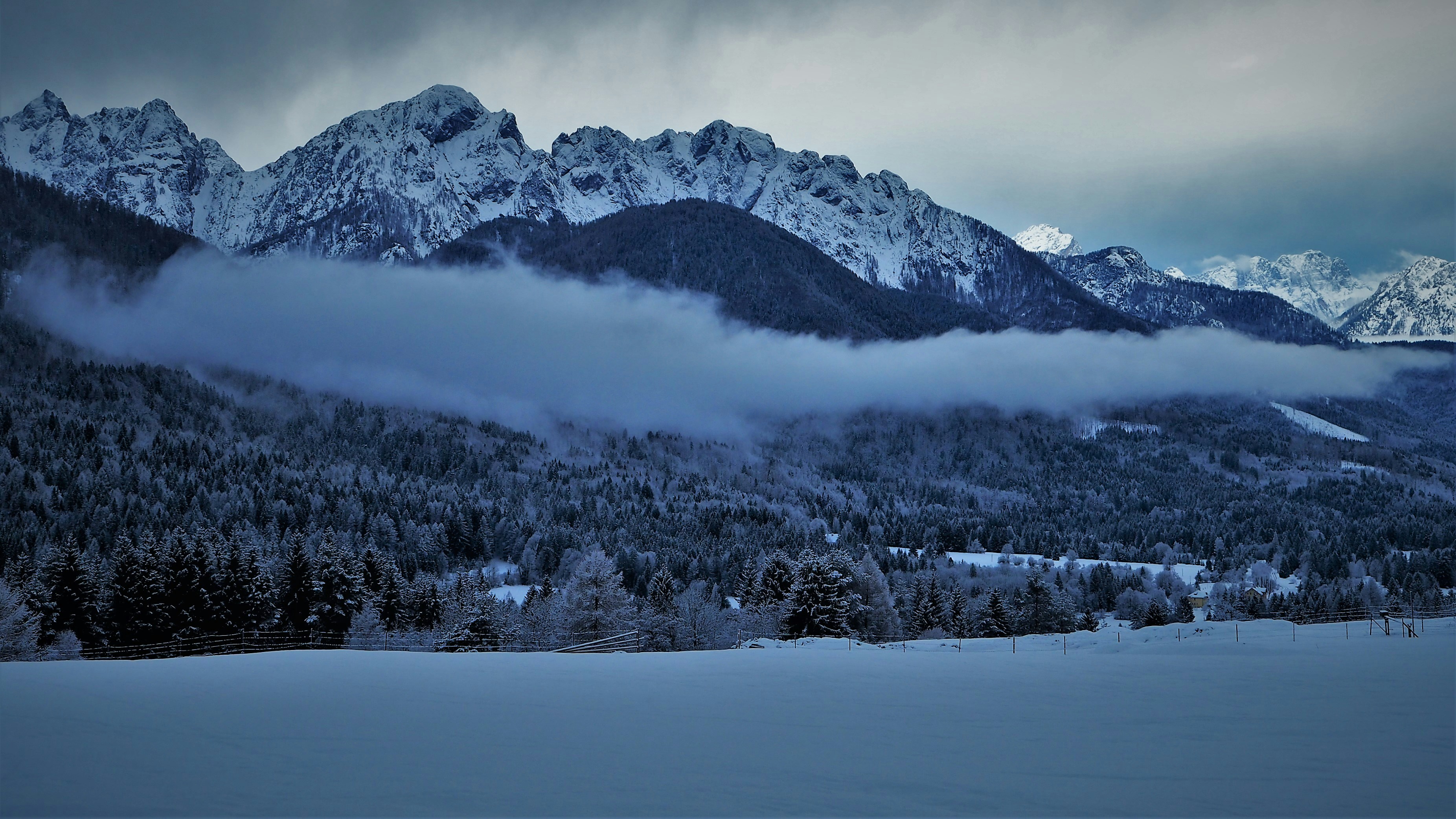 Snow-covered mountains loom over a tranquil valley, with low-hanging clouds weaving through the landscape. The scene captures the serene essence of winter.