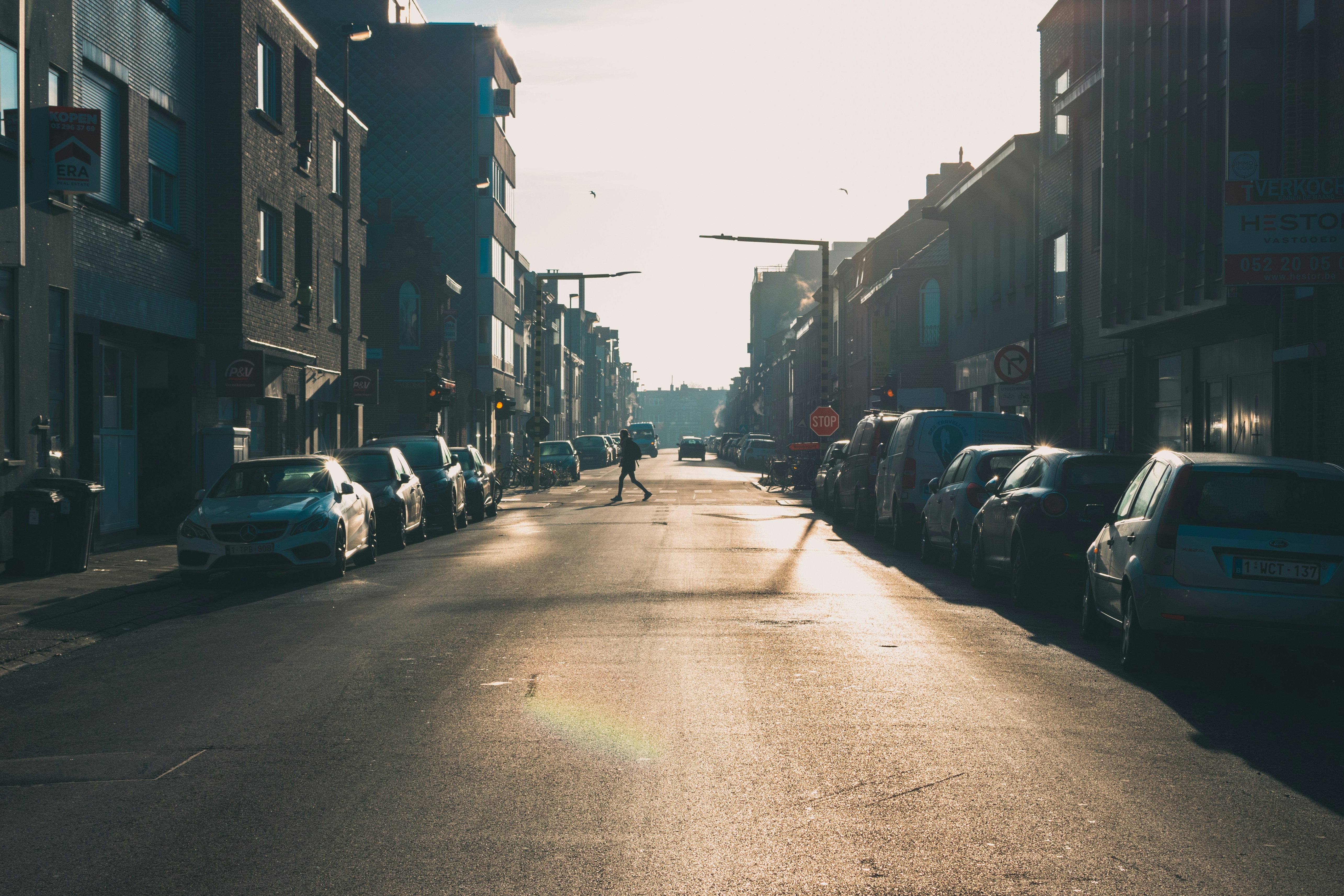 Sunlit street scene with a solitary figure walking amidst parked cars, capturing the essence of urban life.