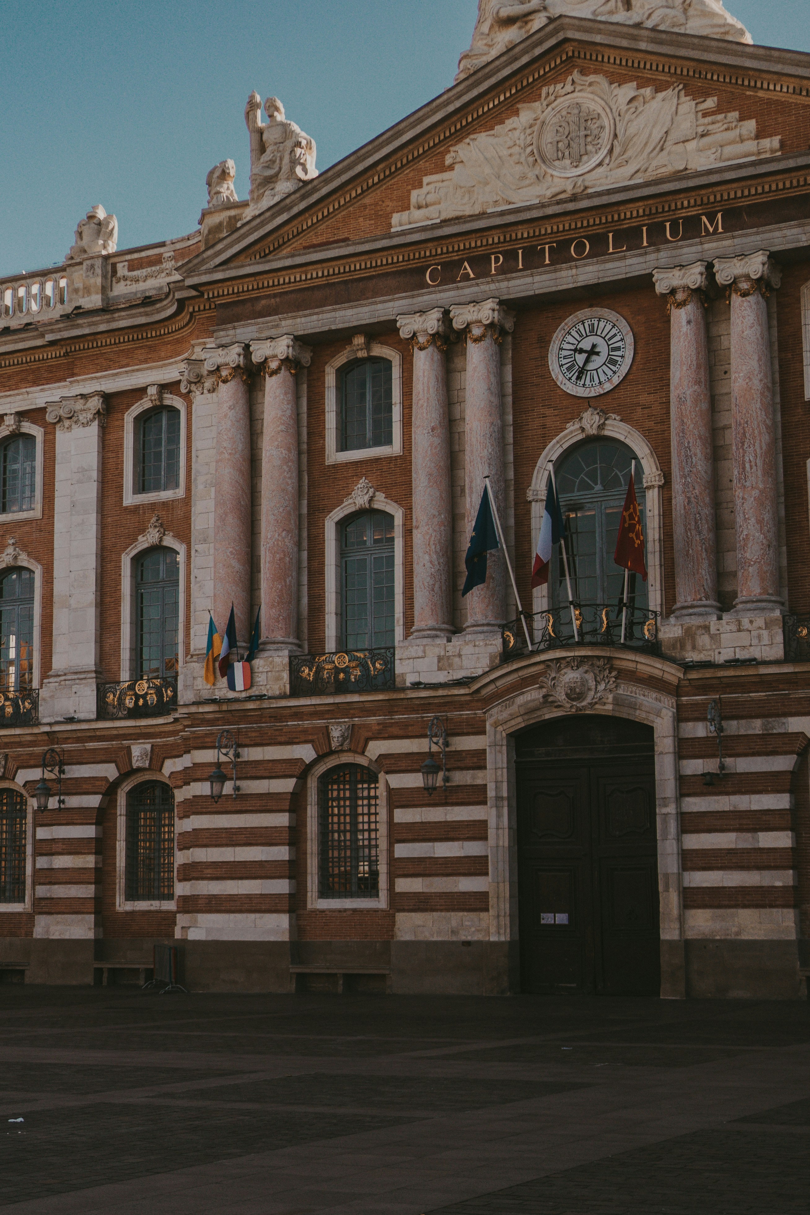 The ornate facade of Capitole in Toulouse showcases classical architecture with vibrant flags and intricate details. The clock adds a touch of timeless elegance.