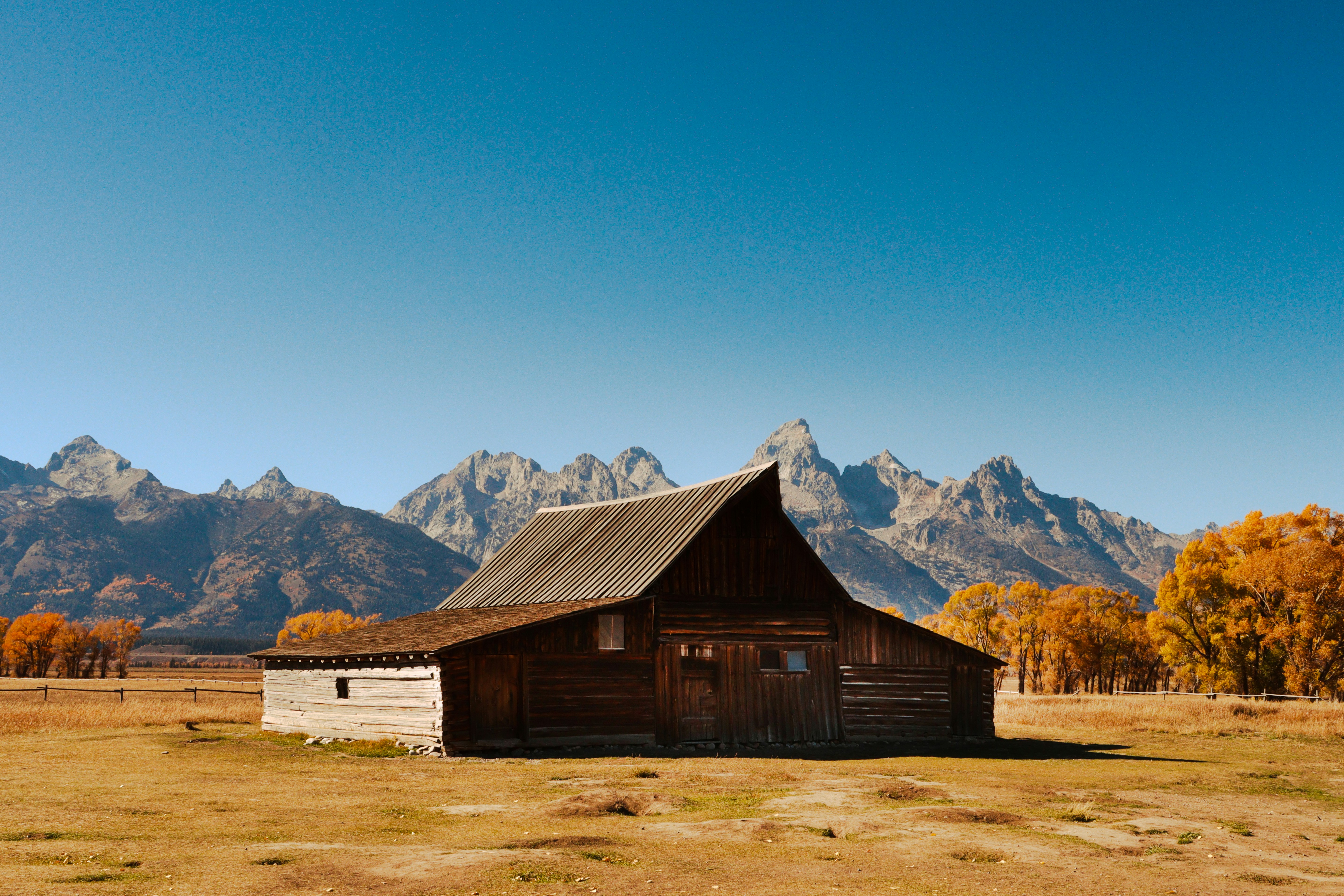 Weathered barn stands in a sprawling field with towering mountains in the background, framed by vibrant autumn foliage.