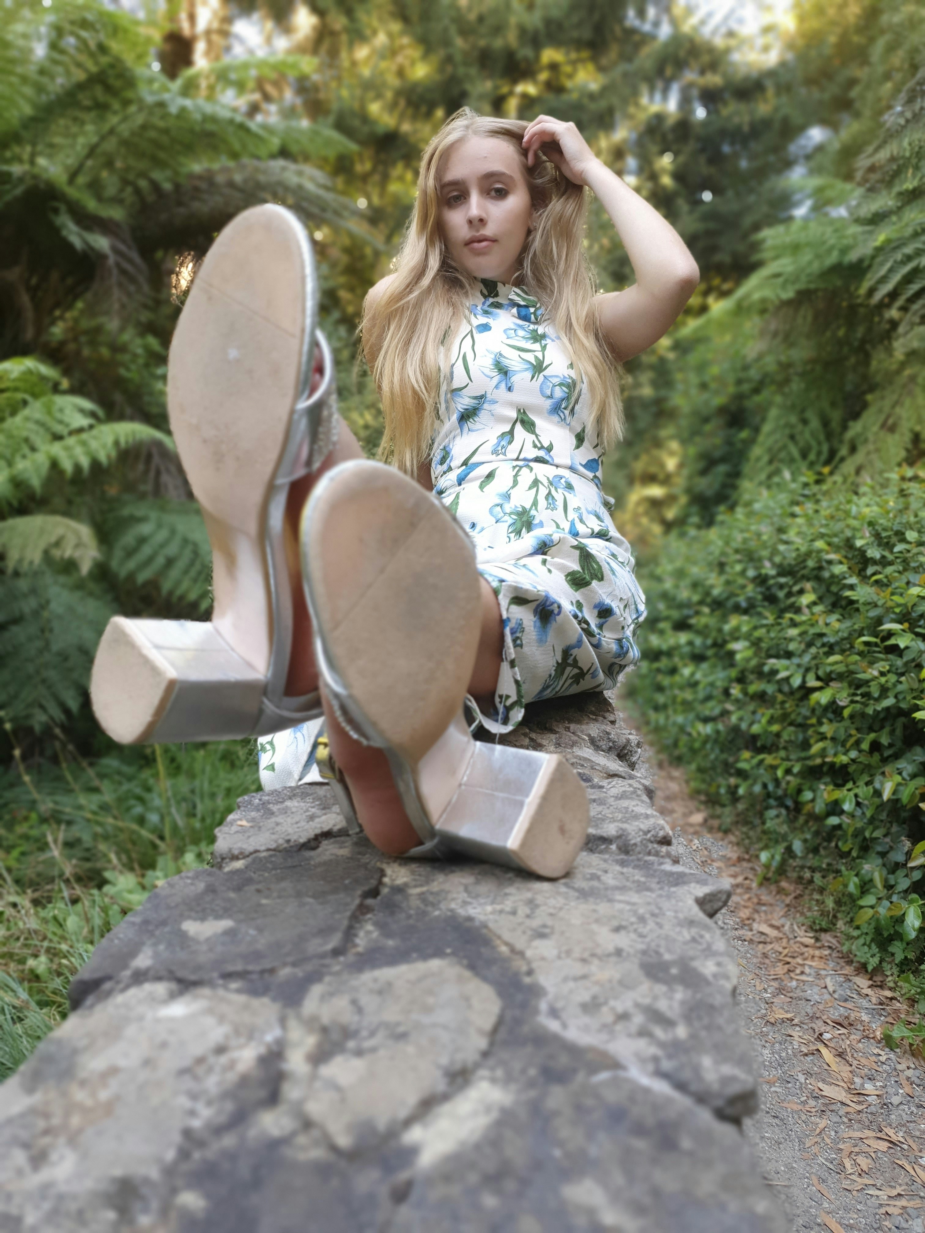 A young woman in a floral dress poses casually on a stone wall, her feet prominently displayed as she enjoys a moment in a lush, green environment.