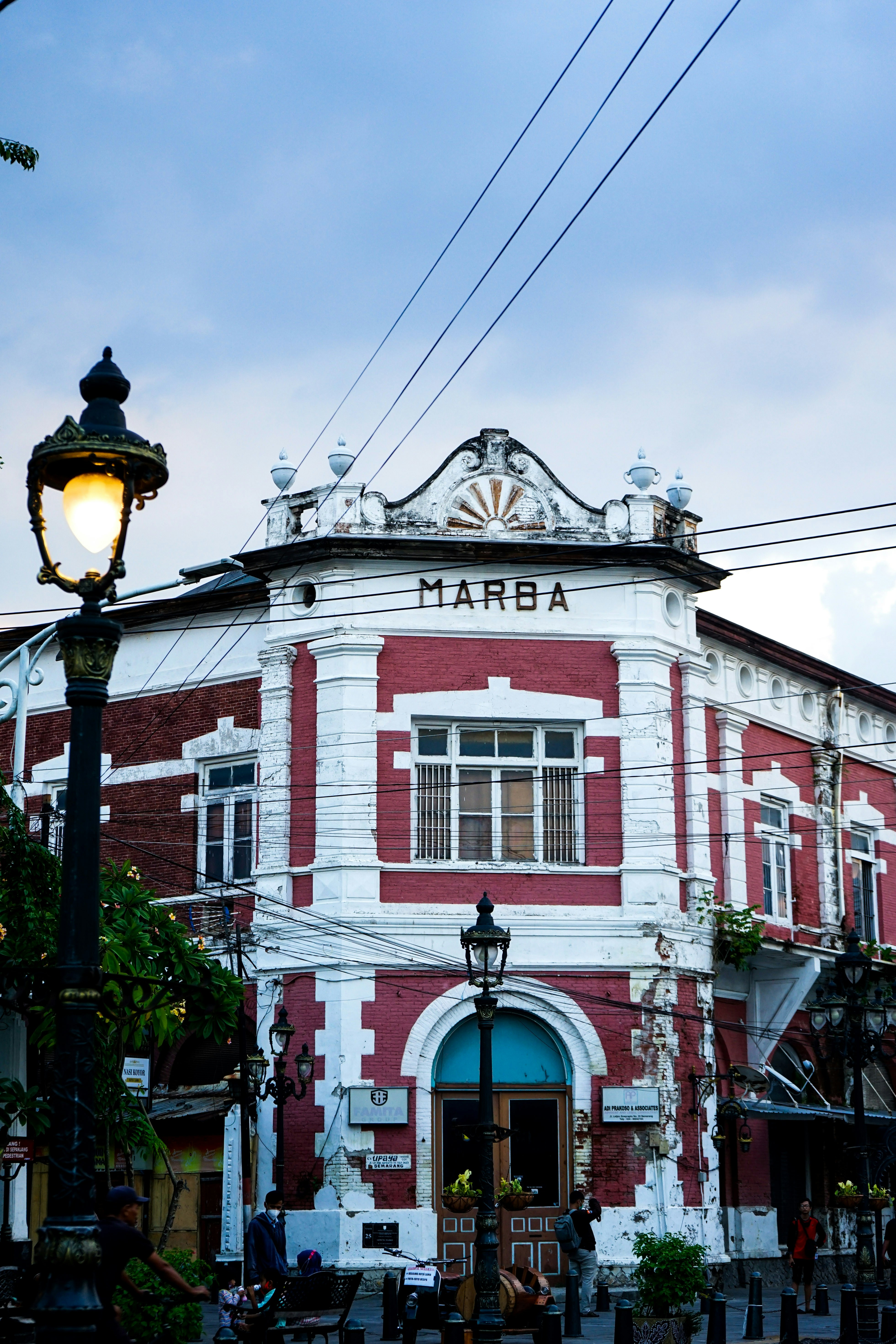 Historic Marba building adorned with intricate architecture, framed by street lamps and lush greenery. The warm glow contrasts with the evening sky.