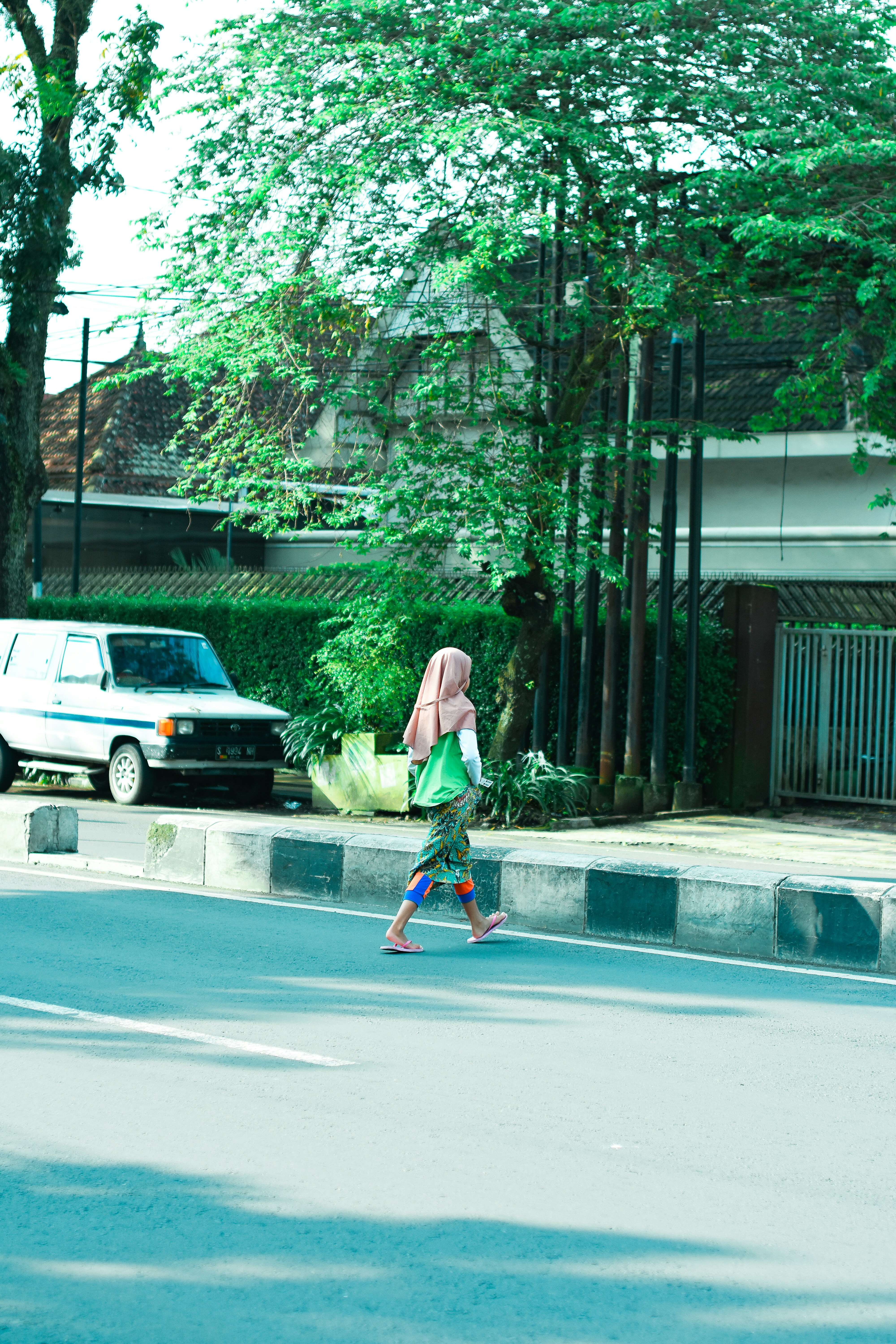 Individual in vibrant clothing walking along a quiet street, surrounded by lush greenery and parked vehicles.