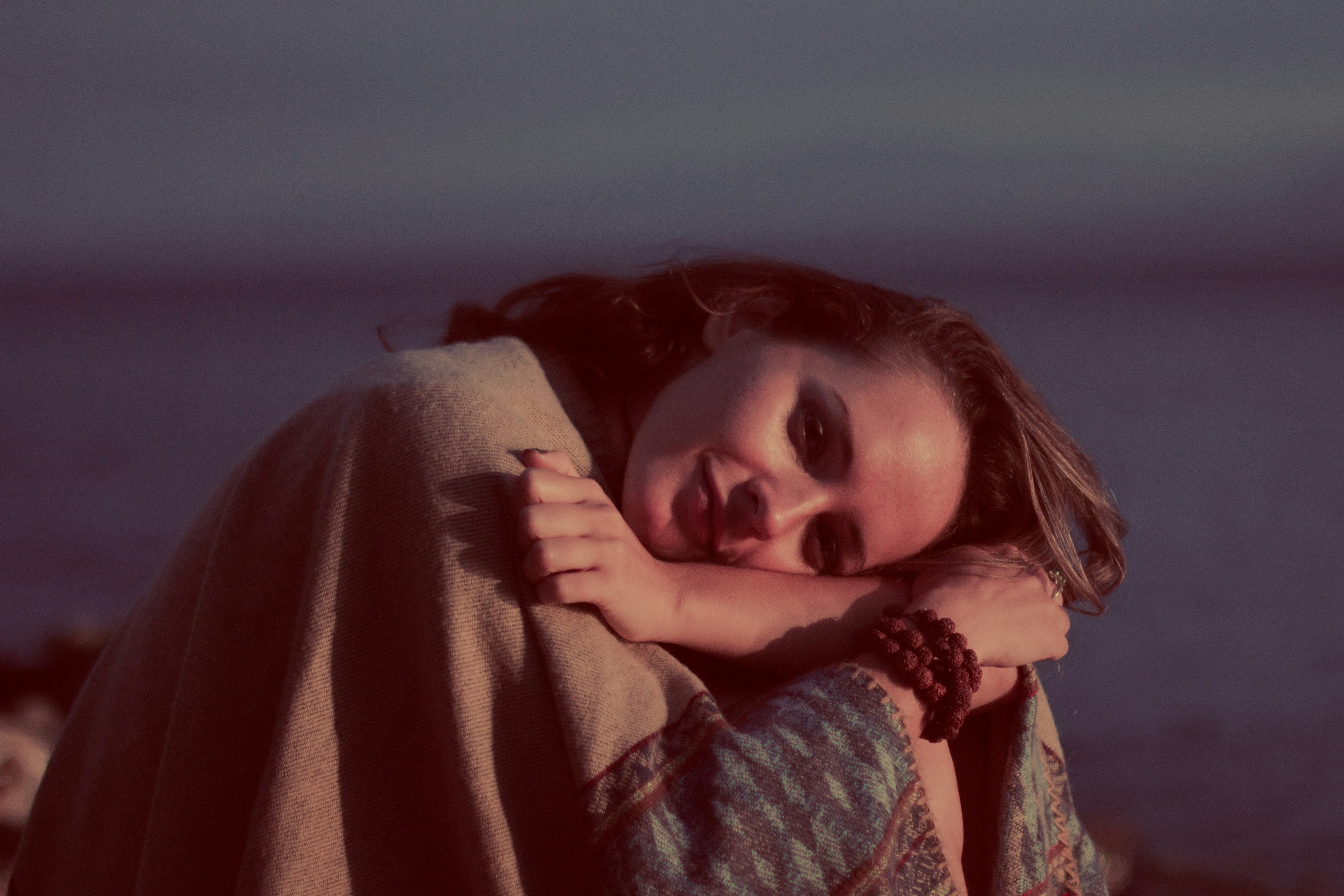 A woman resting her chin on her arms, draped in a cozy blanket, gazes softly at the horizon by the sea.