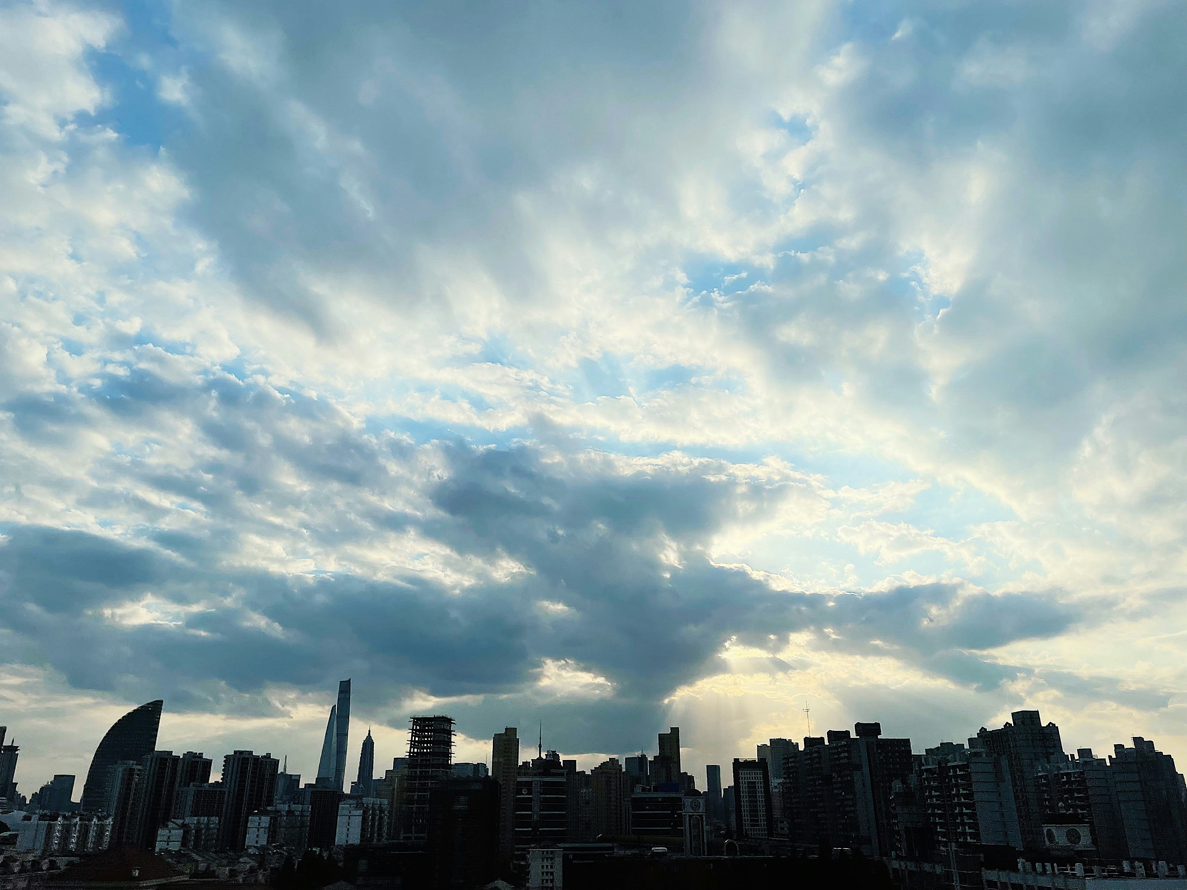 City skyline featuring a mix of modern architecture under a dramatic sky filled with clouds. Light breaks through the overcast, creating a moody atmosphere.