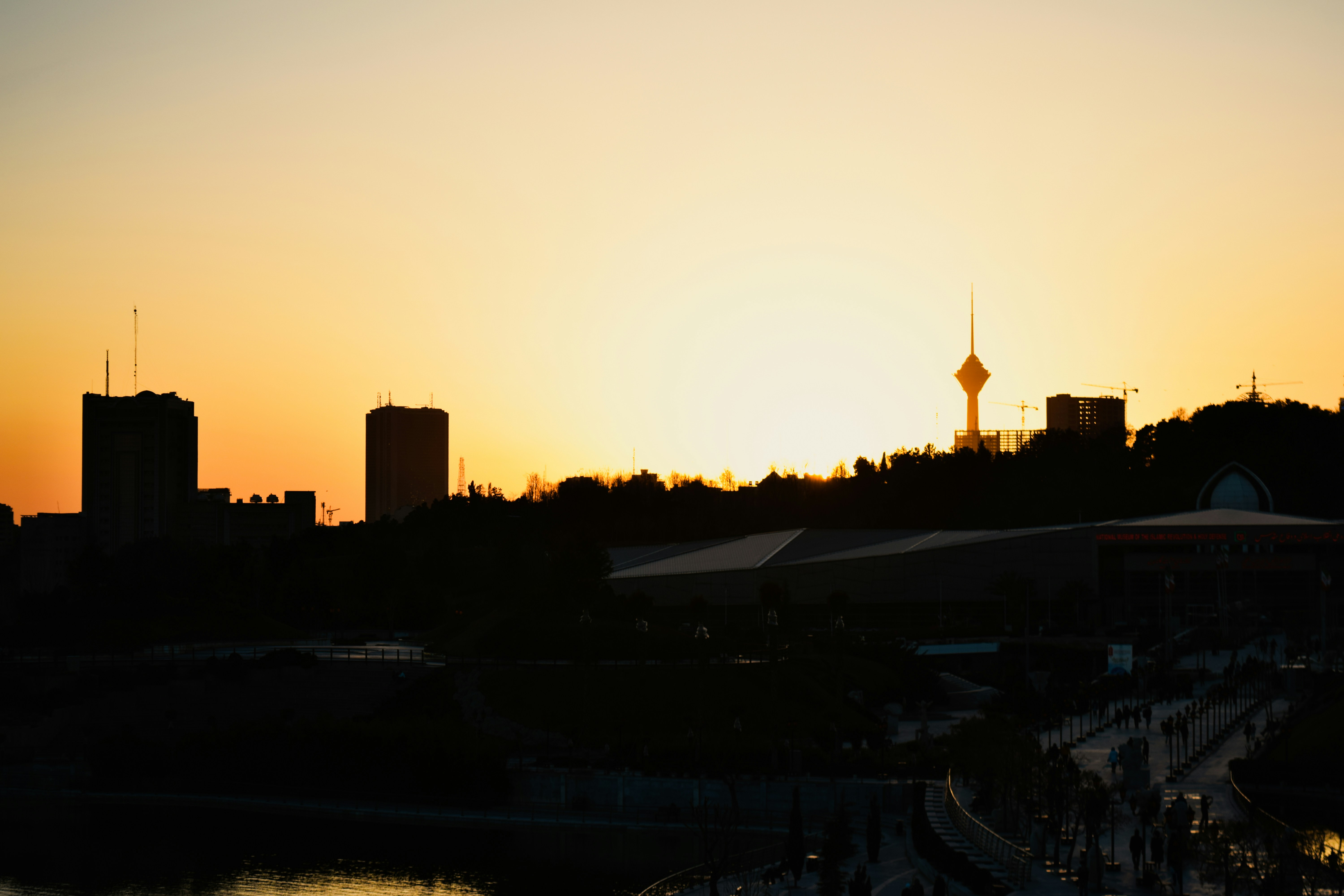 City skyline silhouetted against a vibrant sunset, featuring a prominent tower and bustling waterfront promenade.