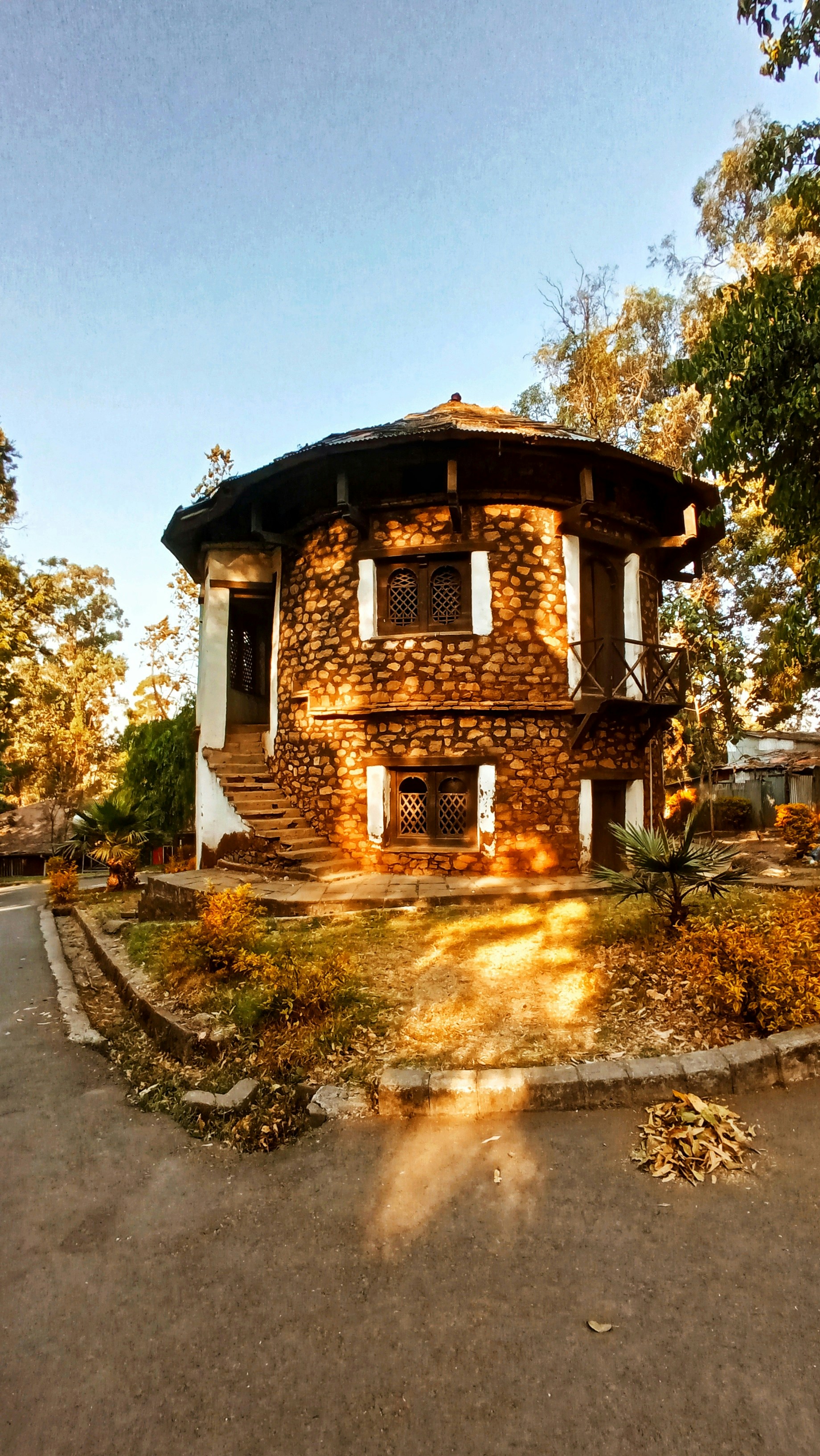 Charming round stone house surrounded by vibrant foliage and sunlight filtering through trees, showcasing a harmonious blend of architecture and nature.