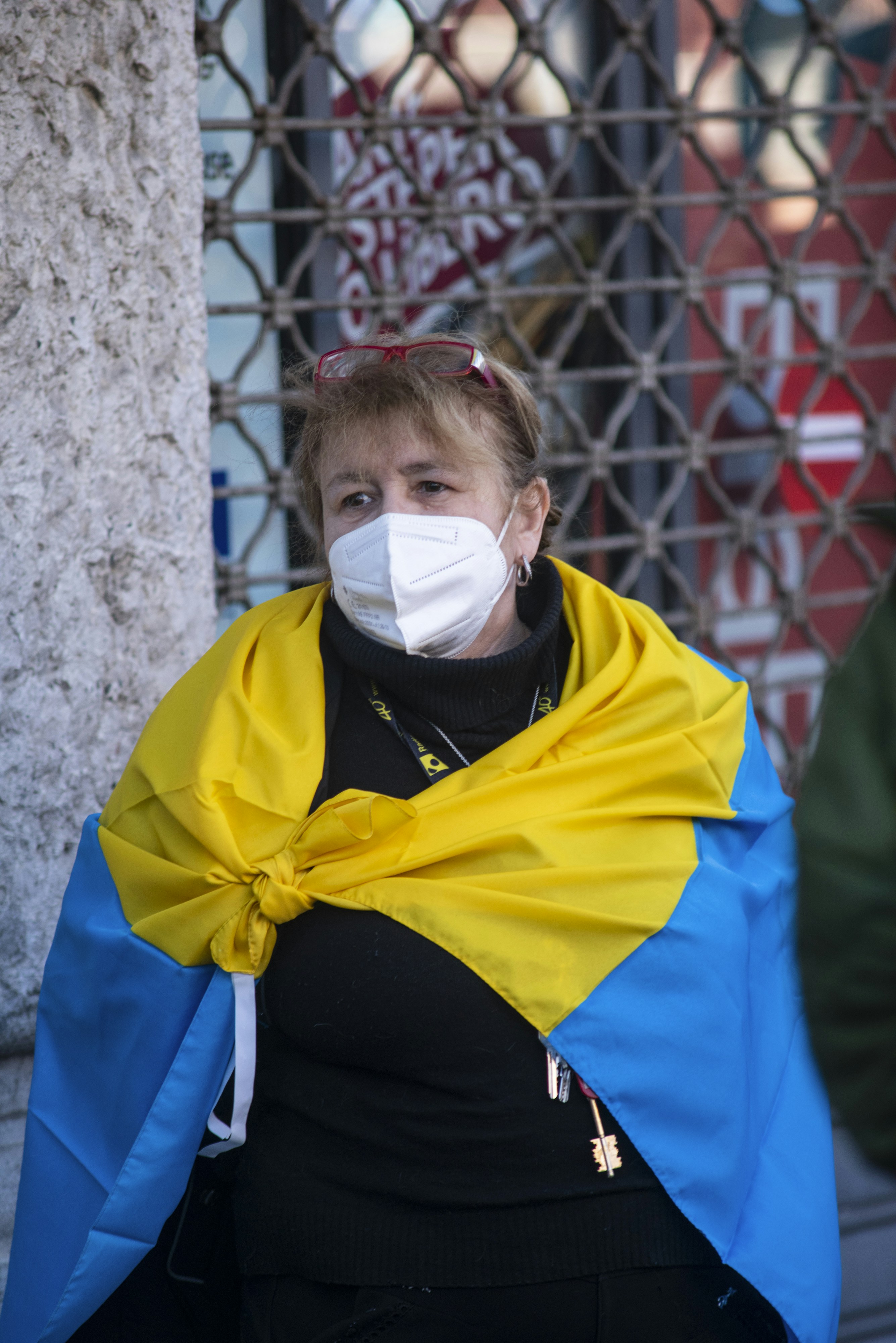 Wrapped in Yellow and Blue: A Masked Observer Beside a Grated Barrier