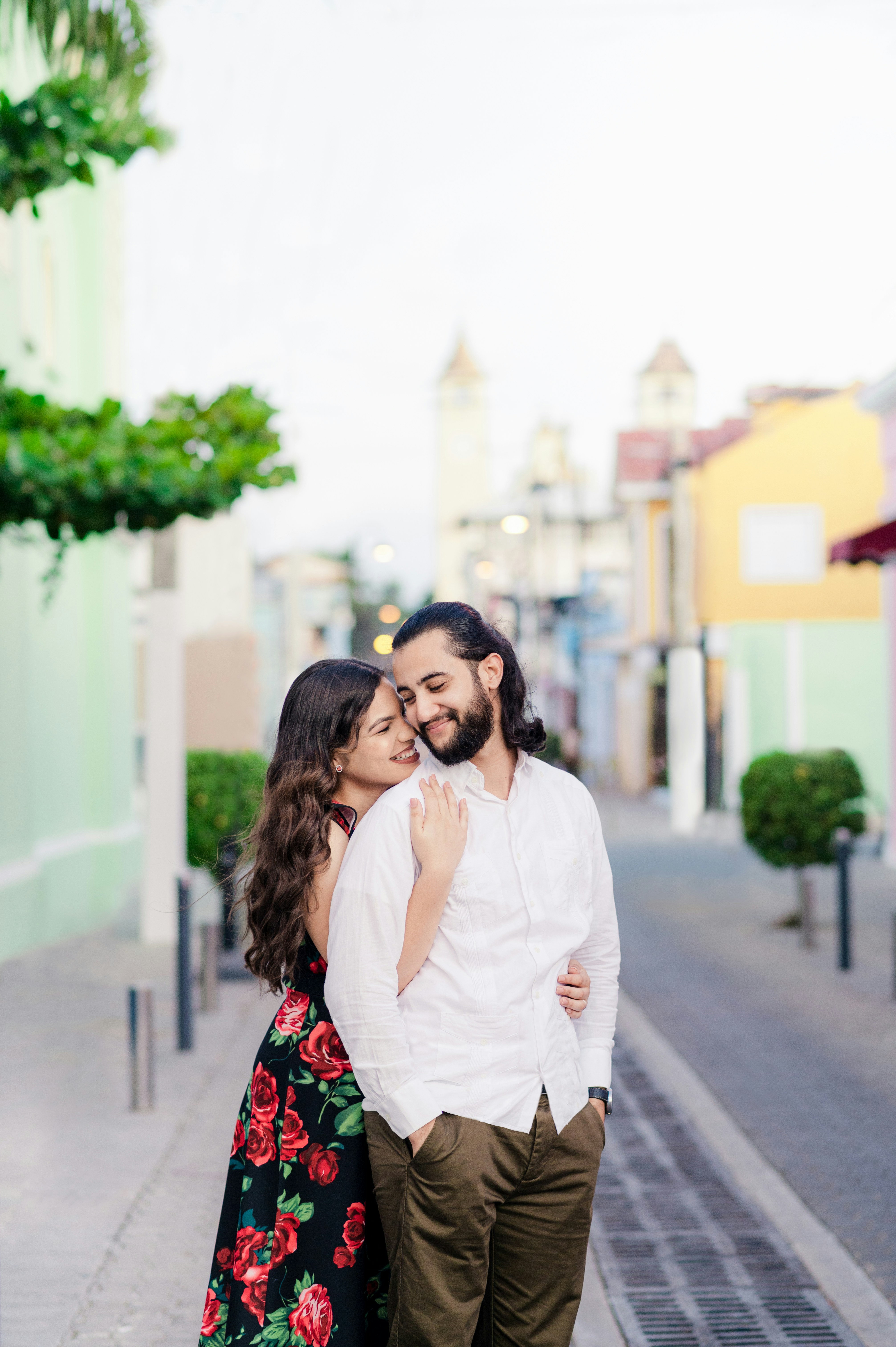 Couple sharing a tender moment on a vibrant street, surrounded by charming pastel buildings. The scene captures a blend of love and local culture.