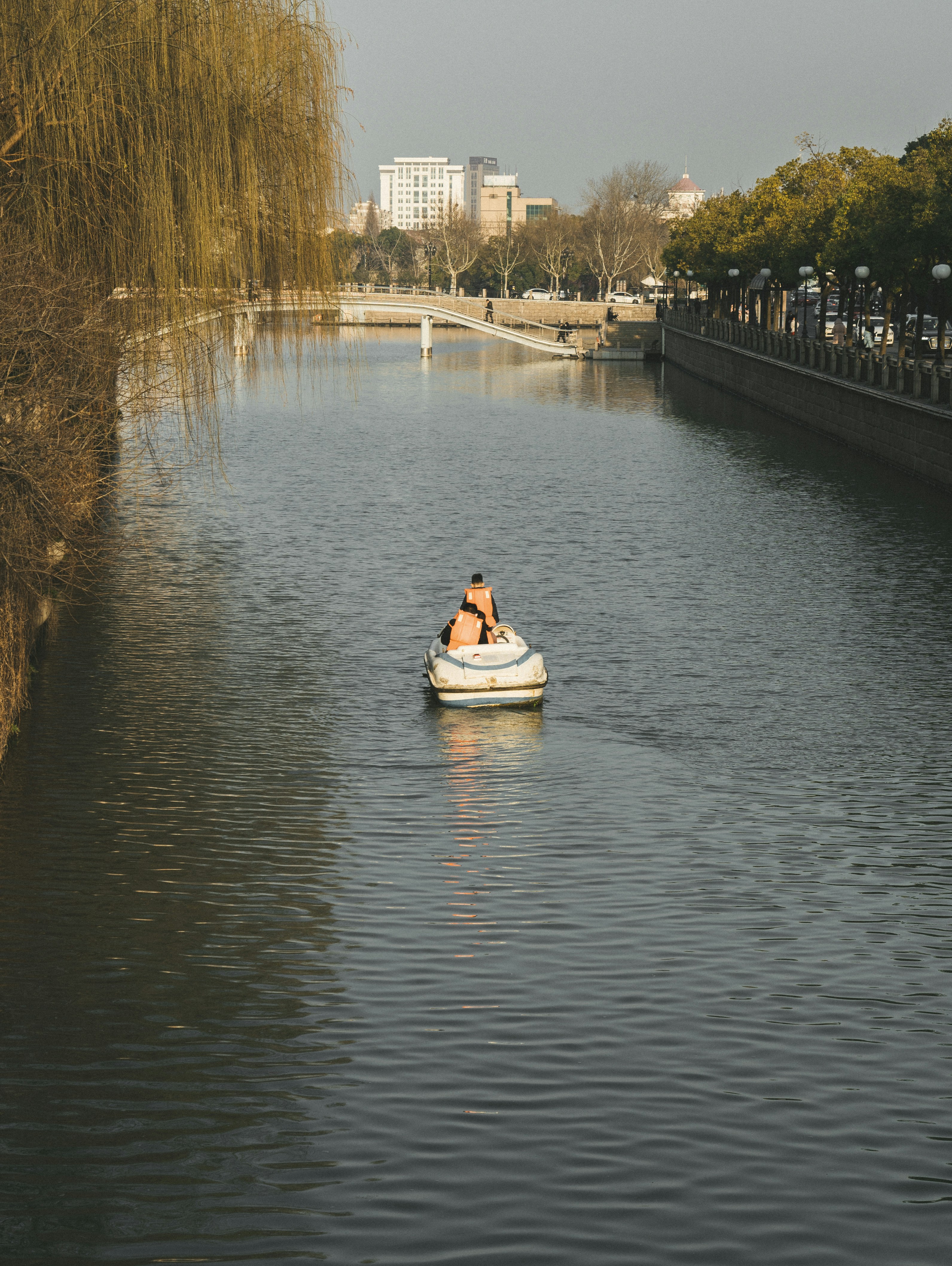 A lone figure in an orange life jacket navigates a small boat along a calm river, surrounded by trees and urban architecture.