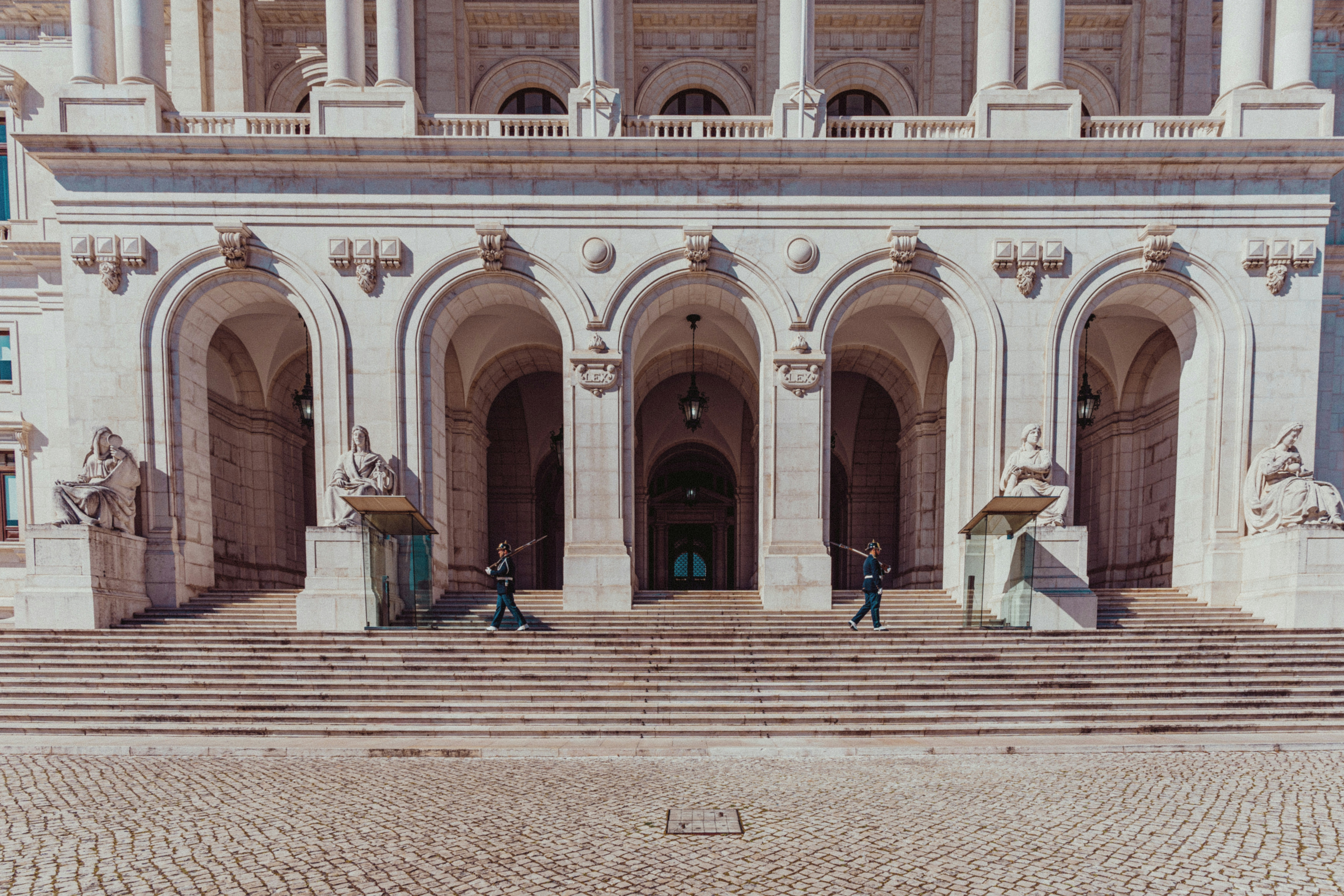 Two guards march in unison down the grand staircase of a neoclassical building, flanked by majestic stone sculptures.