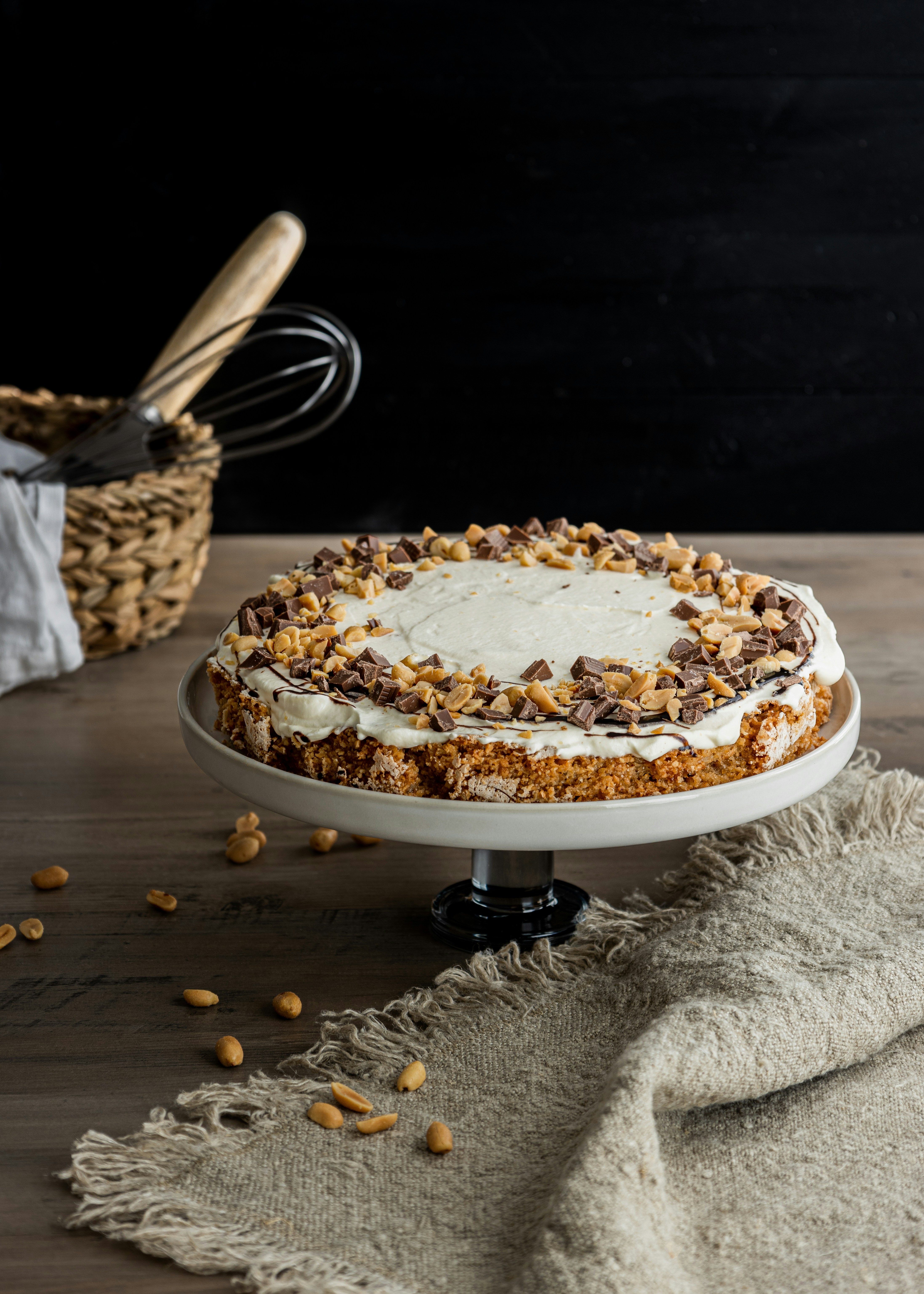 A richly decorated cake topped with chocolate pieces and crushed peanuts, displayed on a white pedestal against a dark background.