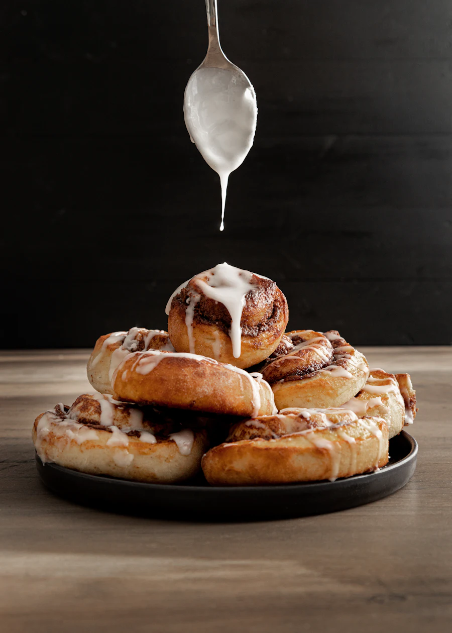 A spoon drips white icing over a stack of cinnamon rolls on a dark plate. The rolls sit on a wooden table against a black background.
