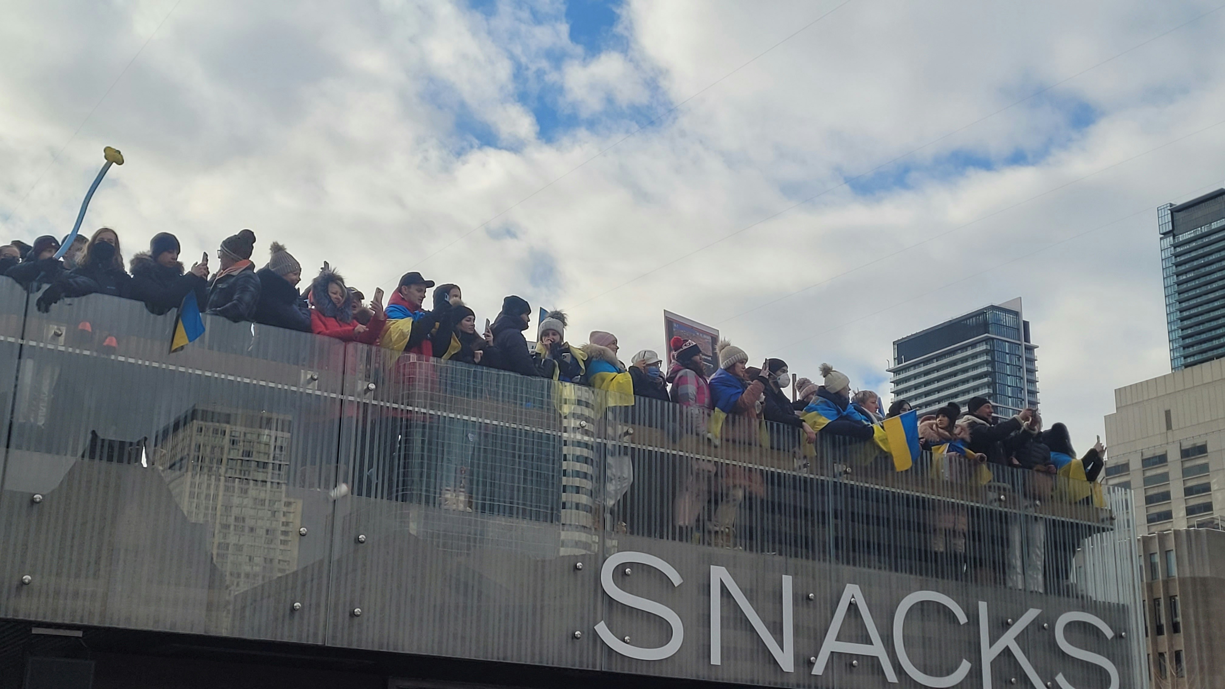 Crowd of people holding flags on a balcony, expressing solidarity during an event. The backdrop features a mix of urban architecture and cloudy skies.