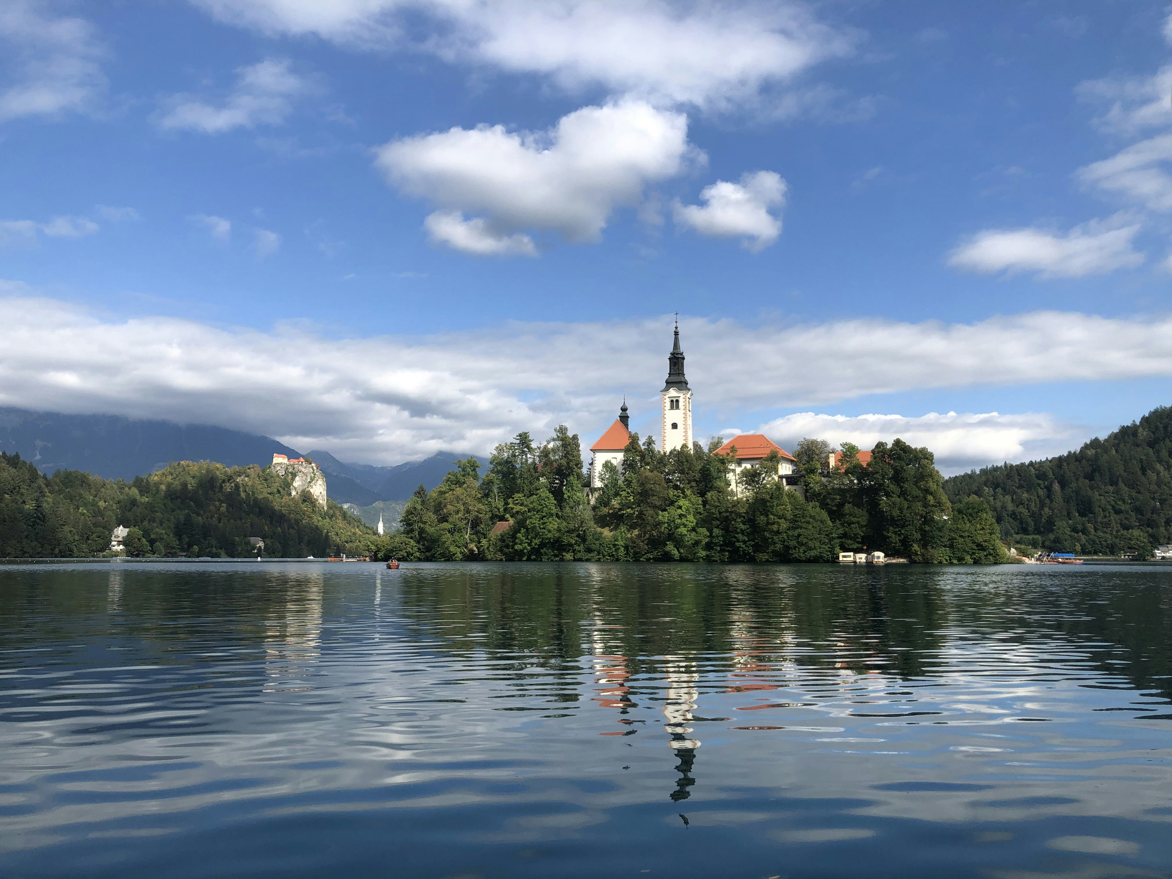 Church on an island surrounded by calm lake waters and lush greenery under a blue sky with scattered clouds.
