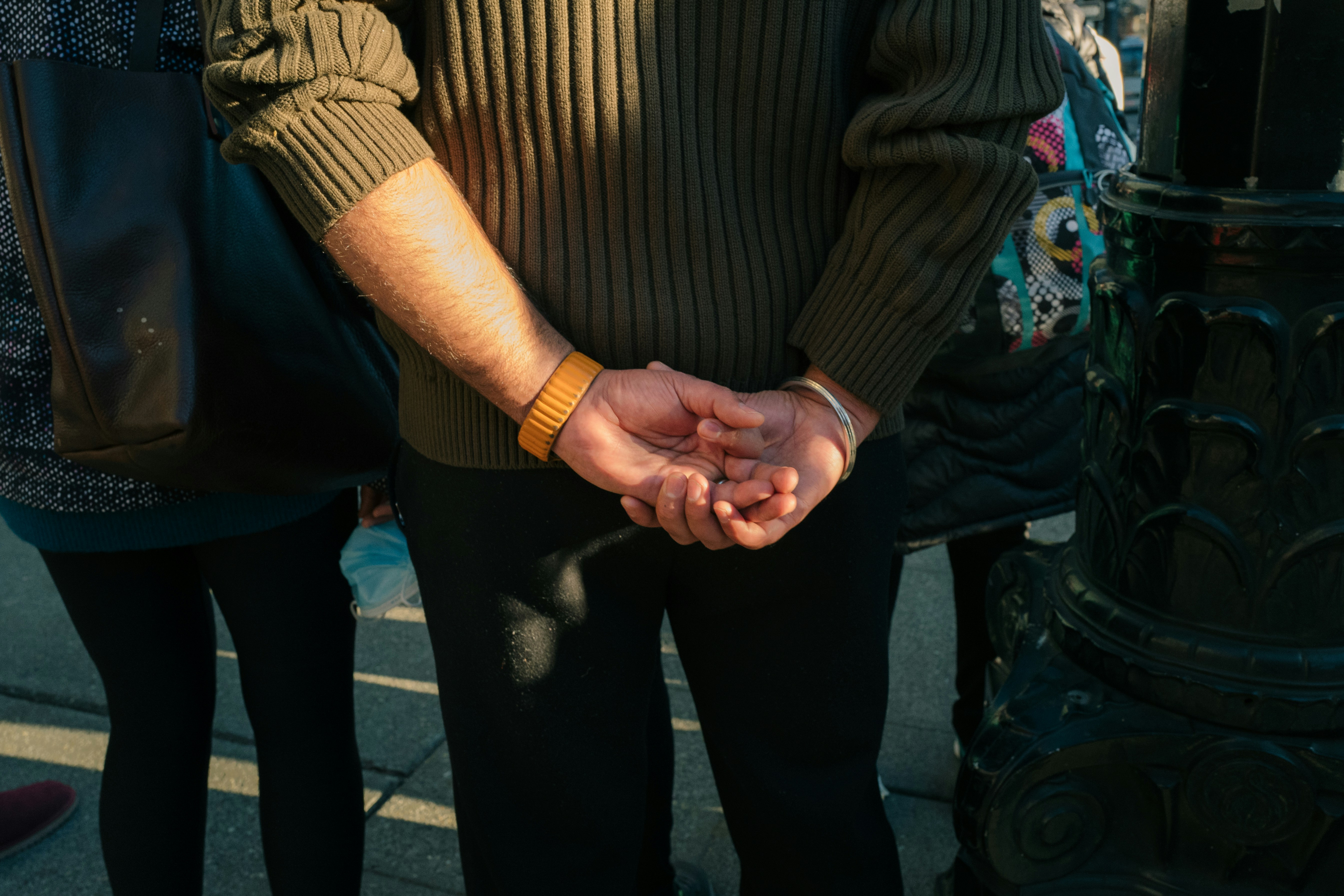 A man stands with his hands clasped behind his back, surrounded by a crowd, suggesting a moment of contemplation or waiting.