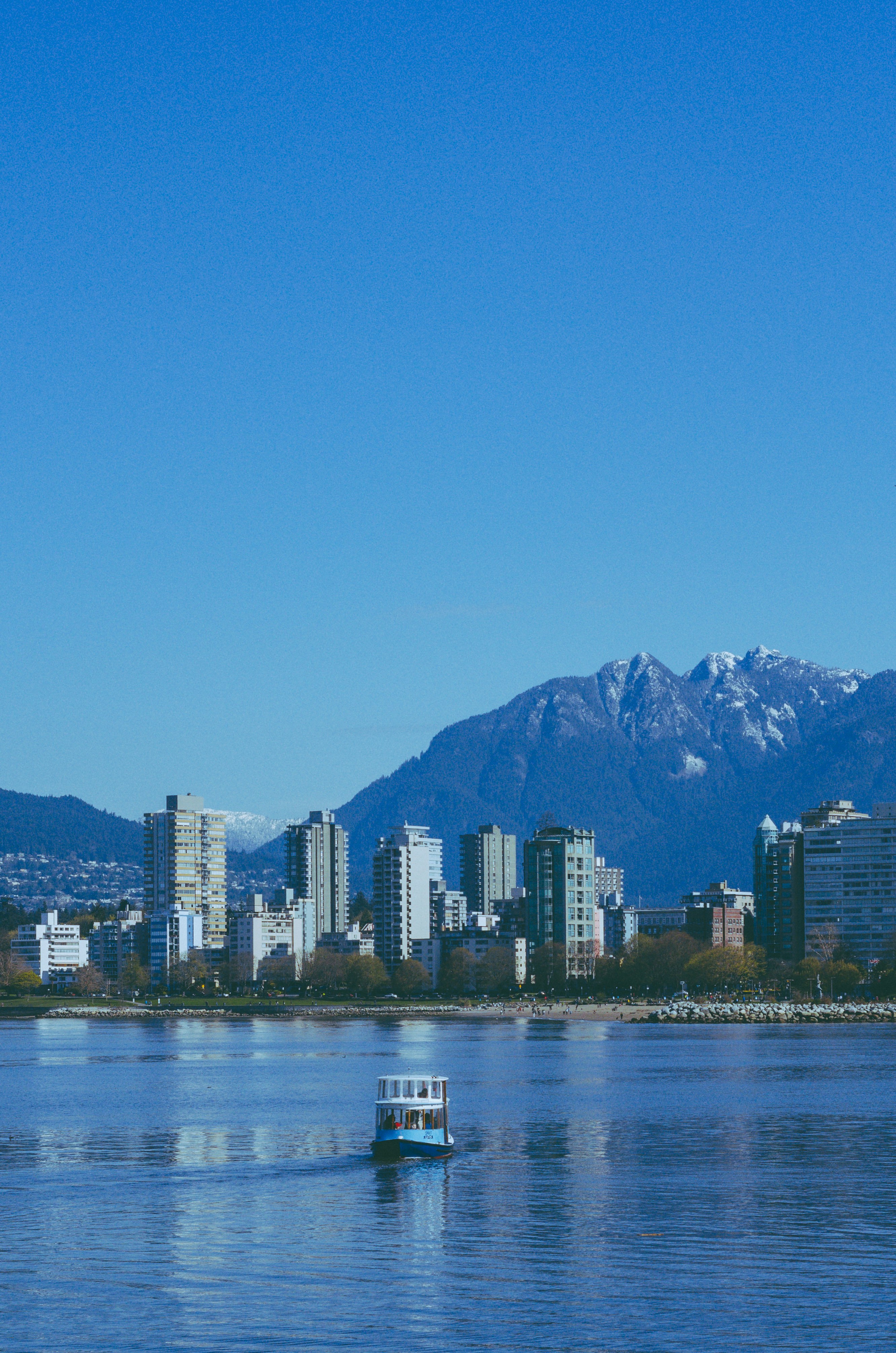 A small boat glides across a tranquil waterway, framed by a city skyline and majestic mountains in the background.