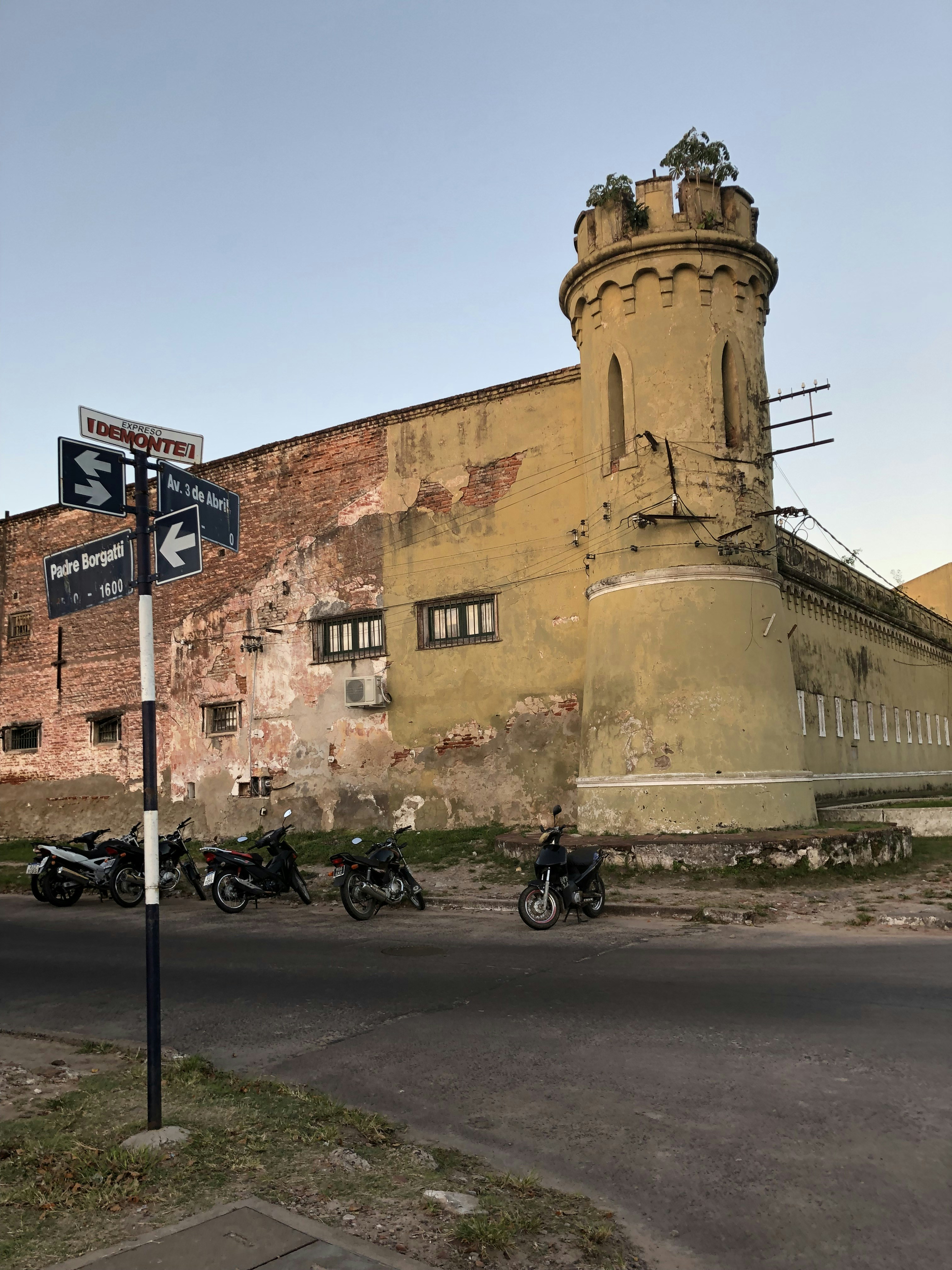 Weathered yellow tower stands against a backdrop of crumbling brick walls, with street signs directing traffic nearby. Motorcycles line the road in the foreground.