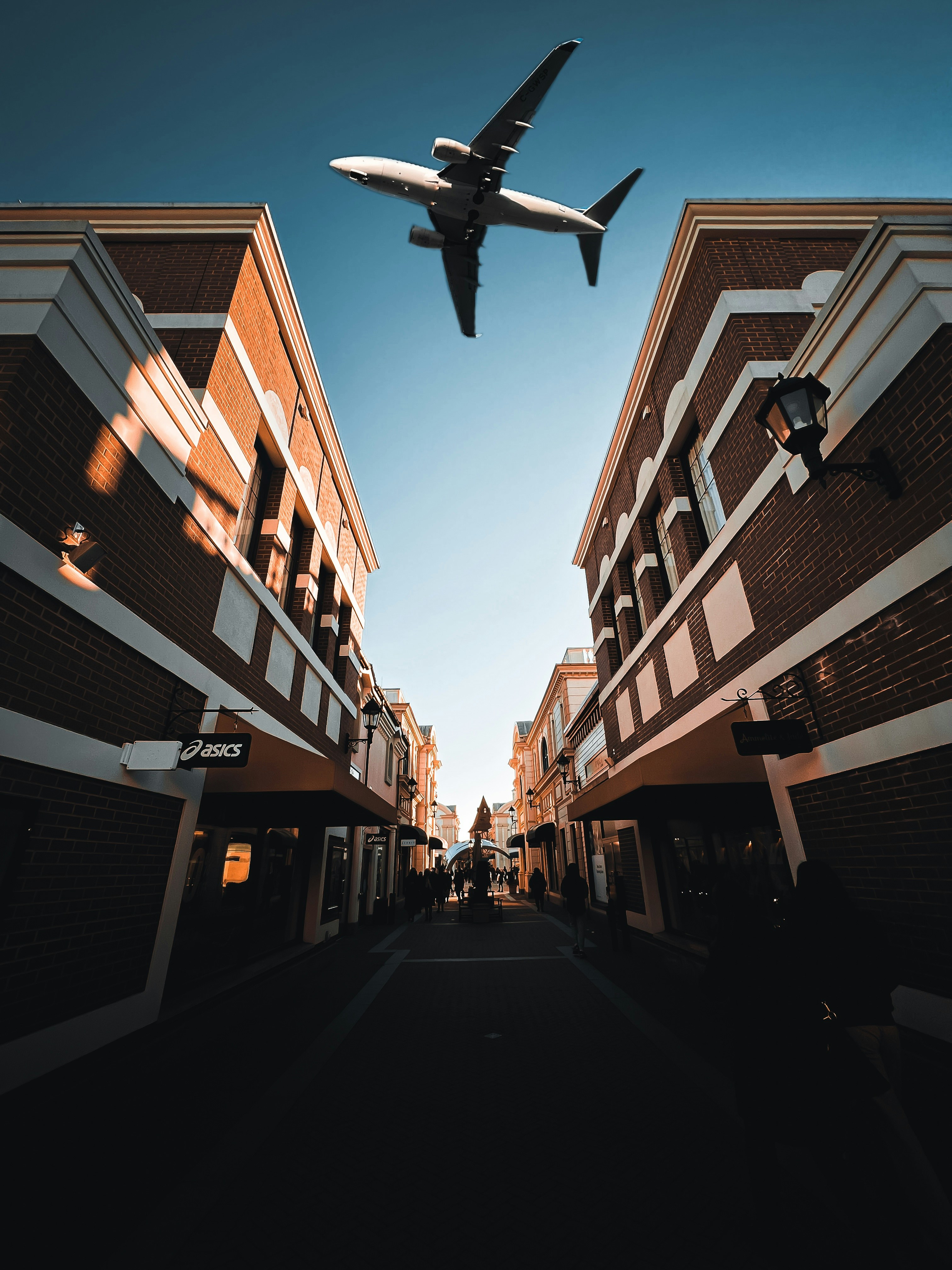 An airplane descends through a narrow alley flanked by brick buildings, highlighting the contrast between urban life and aviation. The clear blue sky serves as a backdrop.