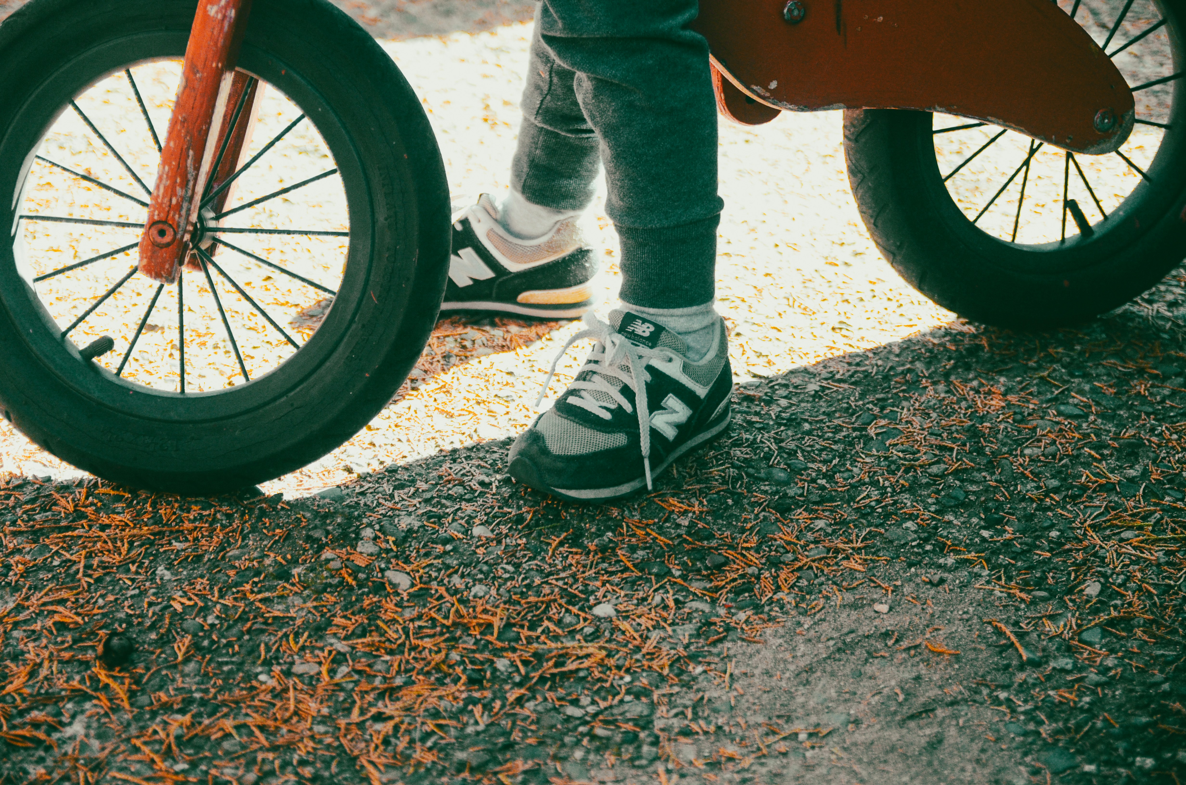 Child's feet clad in New Balance sneakers rest beside a bicycle, with autumn leaves scattered on the ground. The scene captures the essence of childhood exploration.