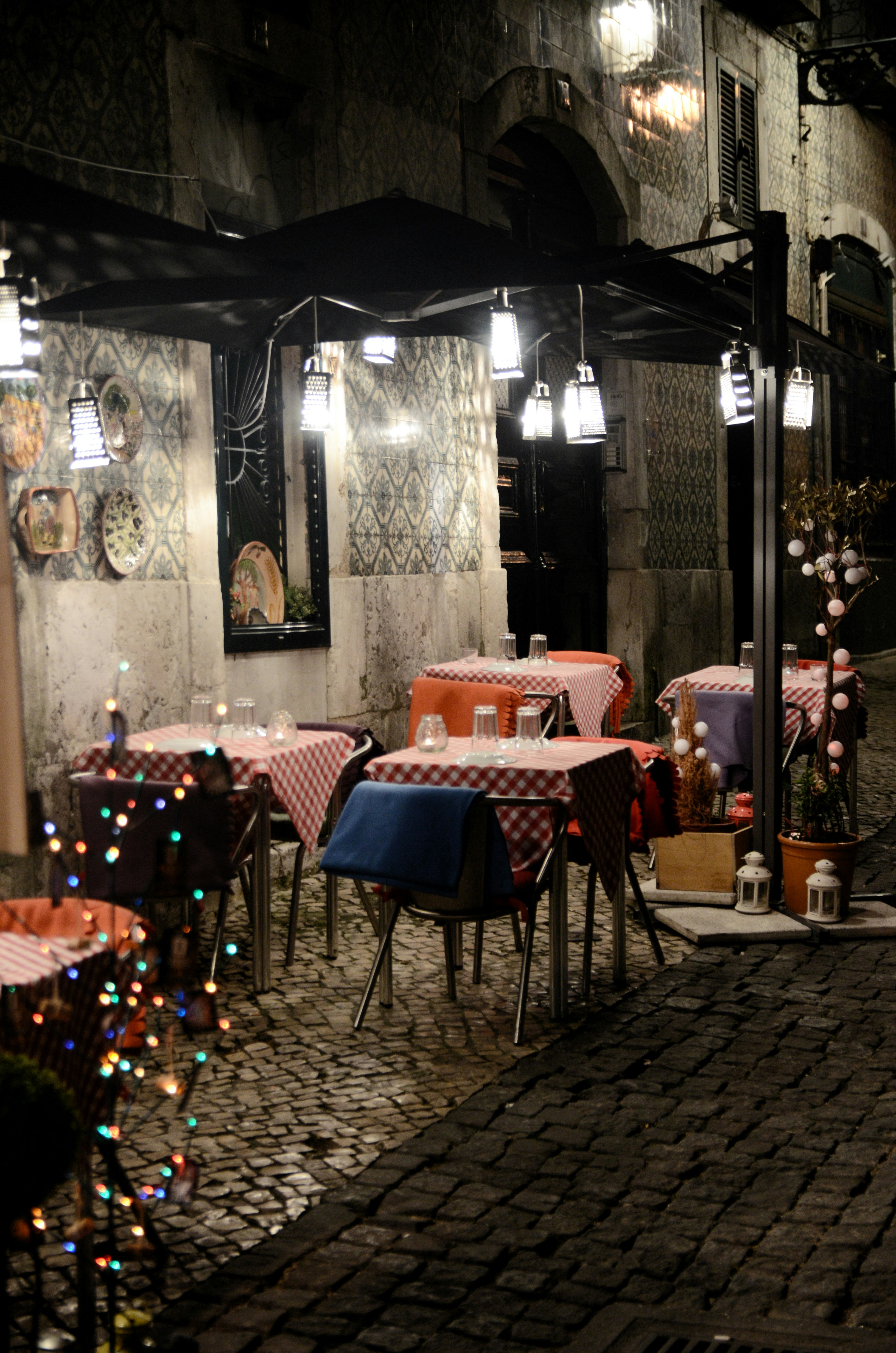 Cozy outdoor dining setup with checkered tablecloths under soft lantern lights, nestled in a cobblestone street adorned with festive decorations.
