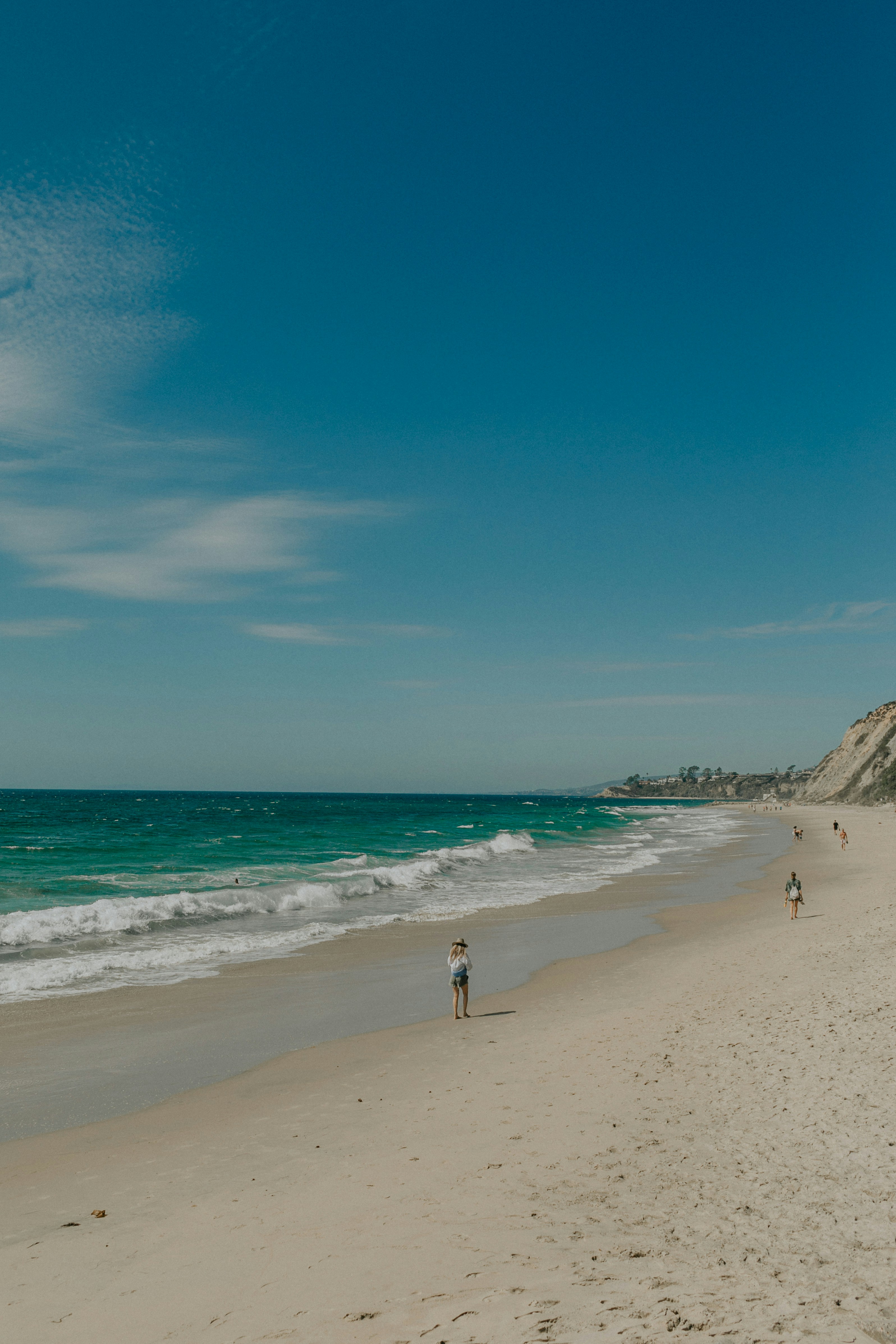 A tranquil beach scene featuring a wide sandy shore and gentle waves, with a few figures enjoying the sunny day. The clear blue sky enhances the peaceful atmosphere.