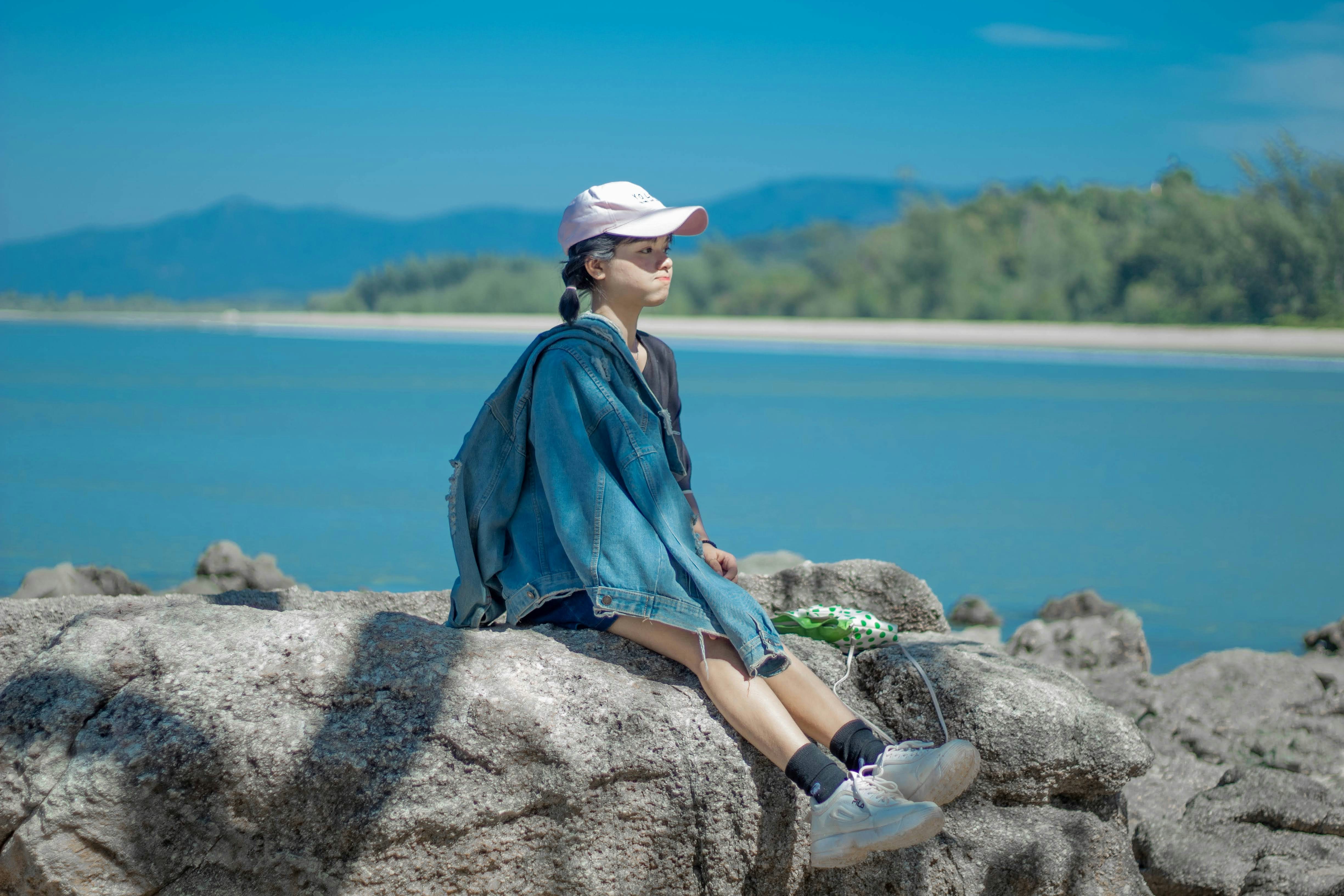 Woman in casual attire sitting on a rock by a tranquil body of water under a clear blue sky.