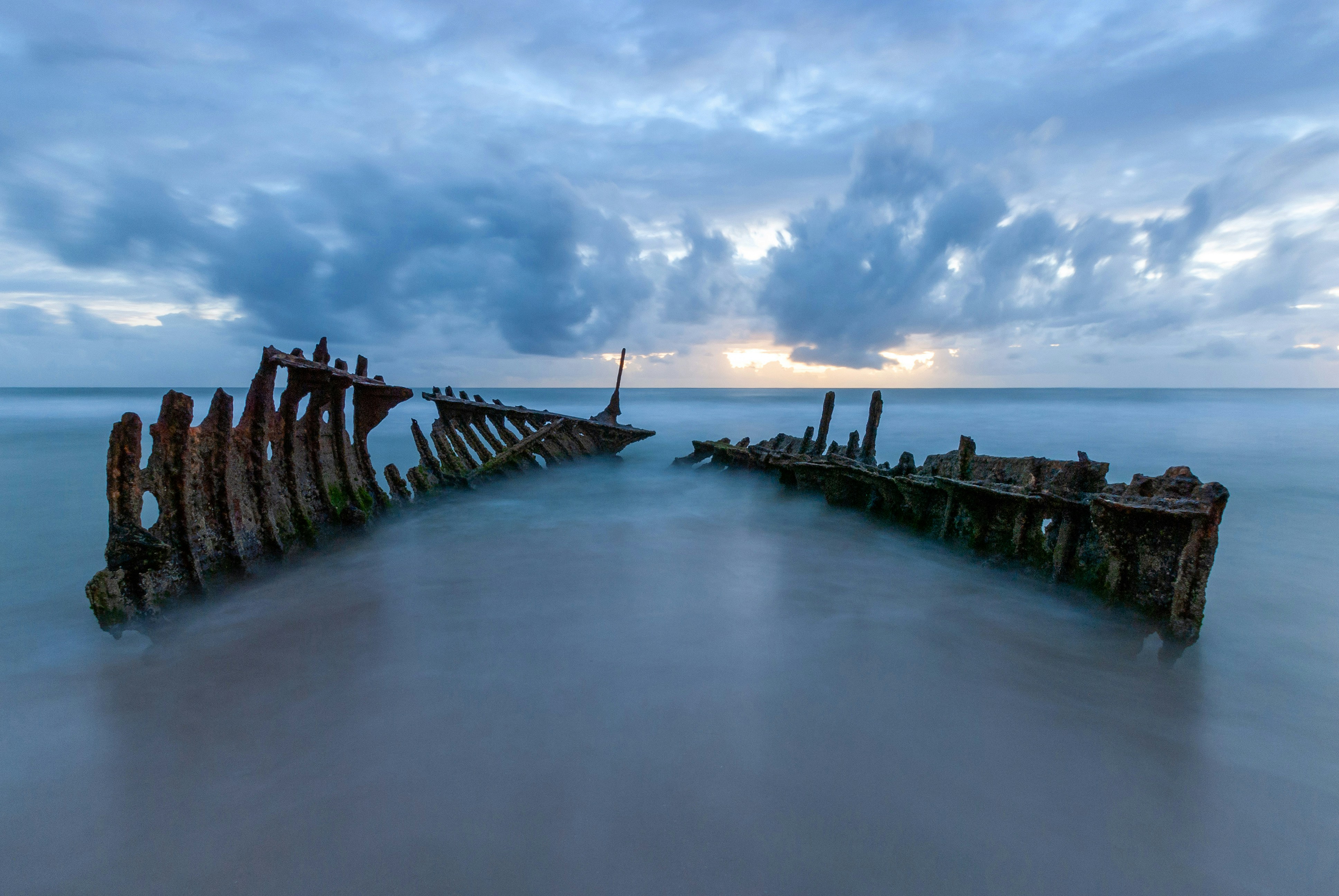 Rusting shipwreck remains emerge from calm ocean waters beneath a dramatic evening sky.