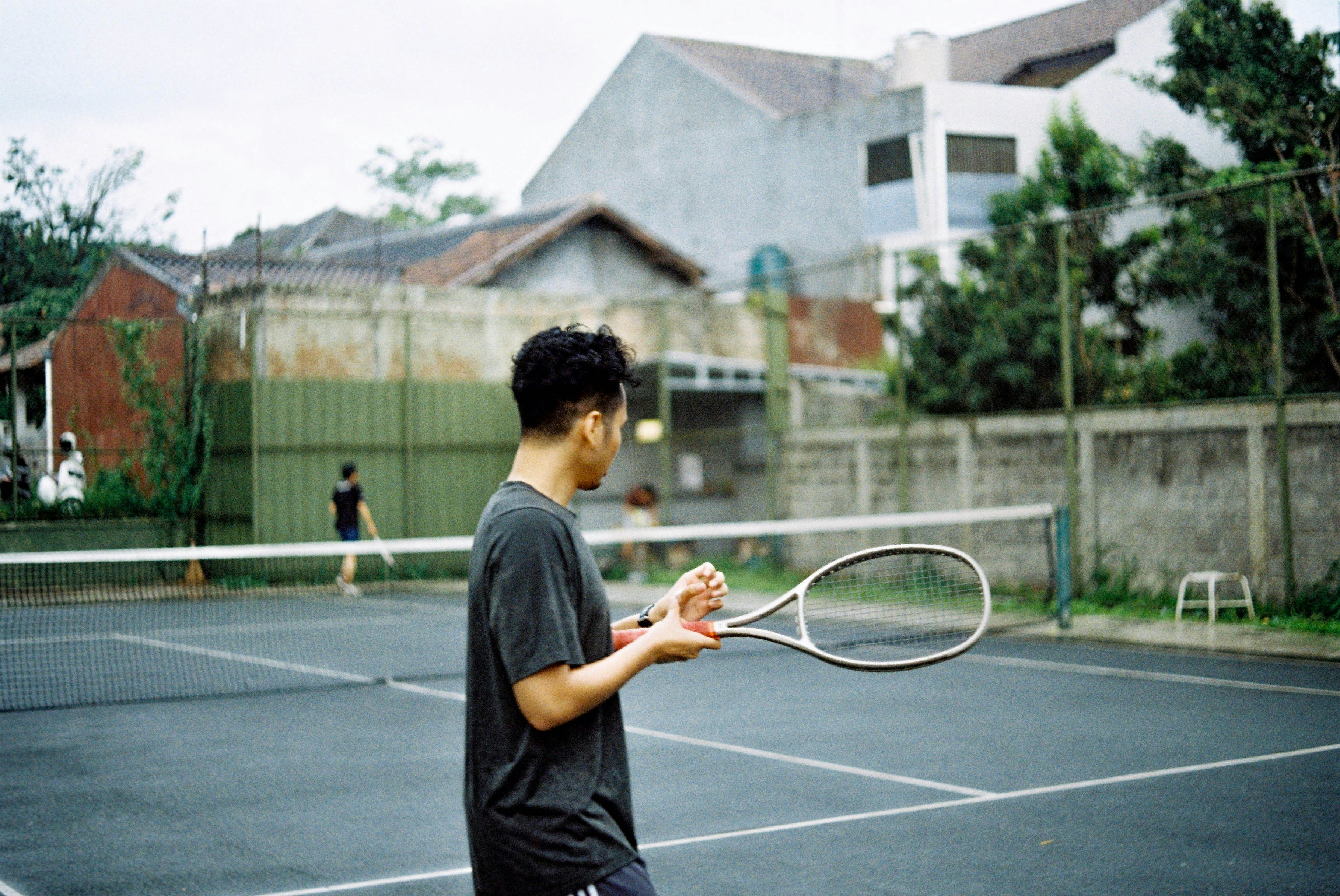 Young man poised with a tennis racket on a court, surrounded by a lively backdrop of players and greenery.