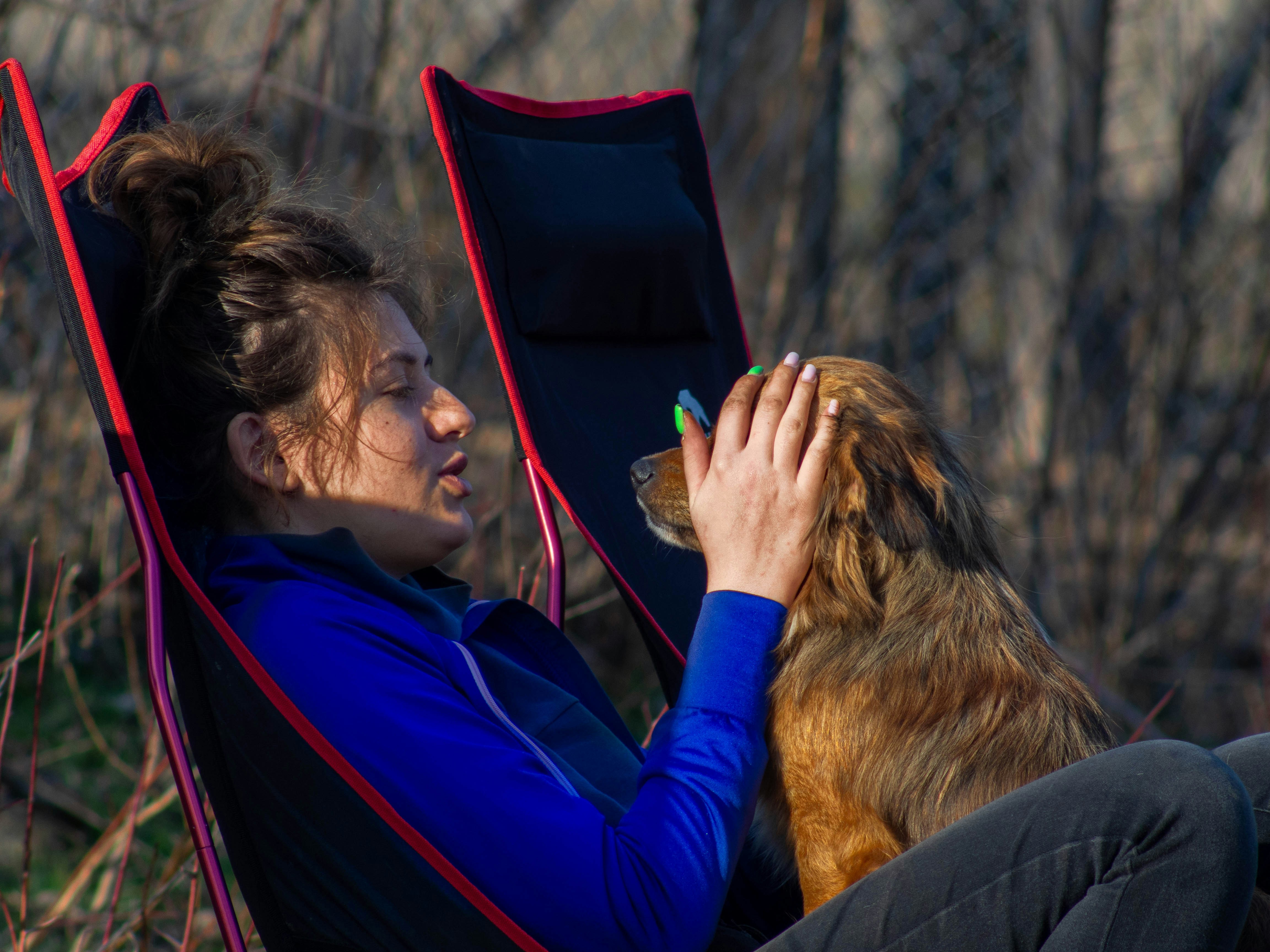 A woman sits in a camping chair, sharing a tender moment with her dog in a natural setting. The scene captures the bond between them.