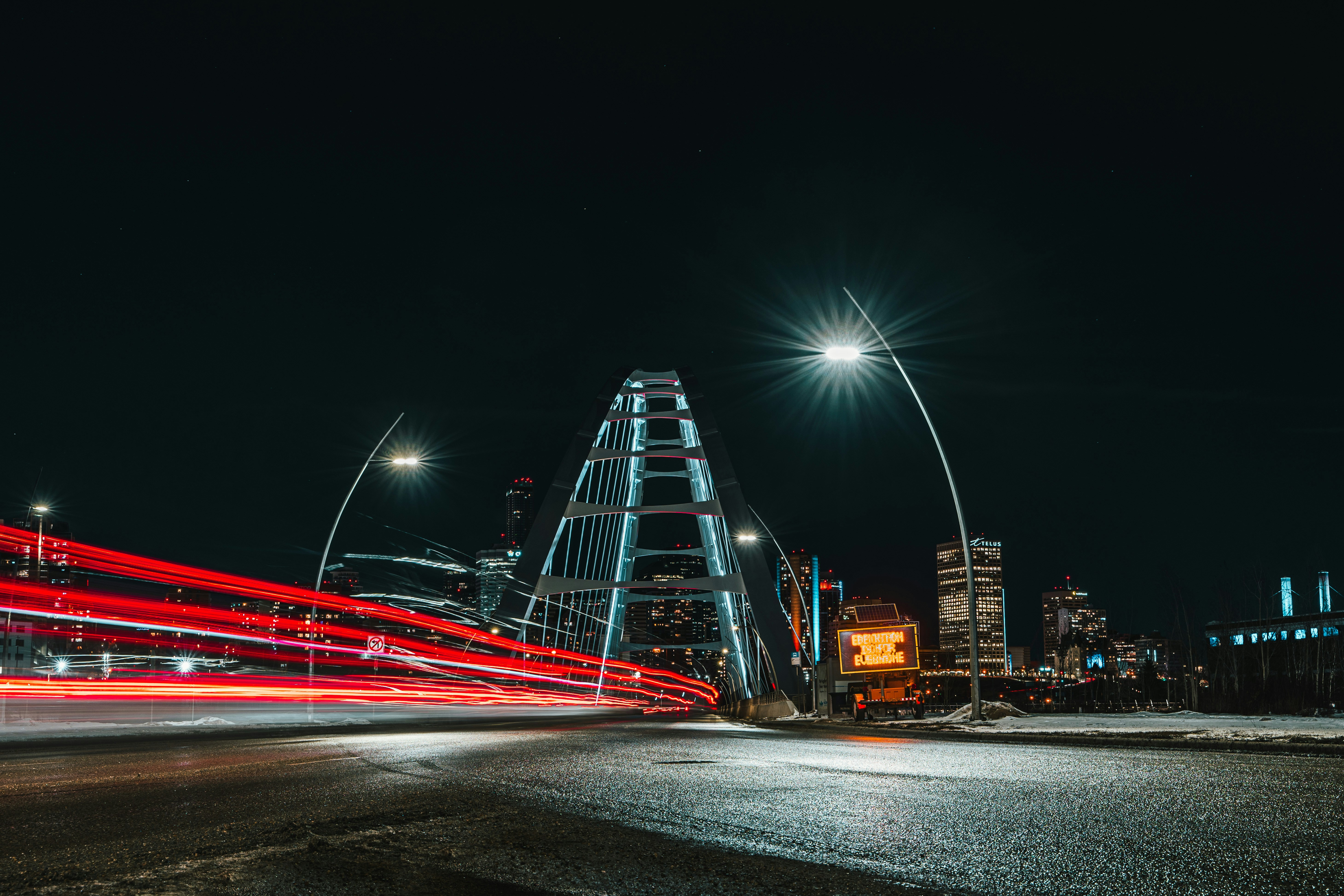 A modern bridge illuminated against a night skyline, with streaks of red light from passing vehicles creating a dynamic contrast. 