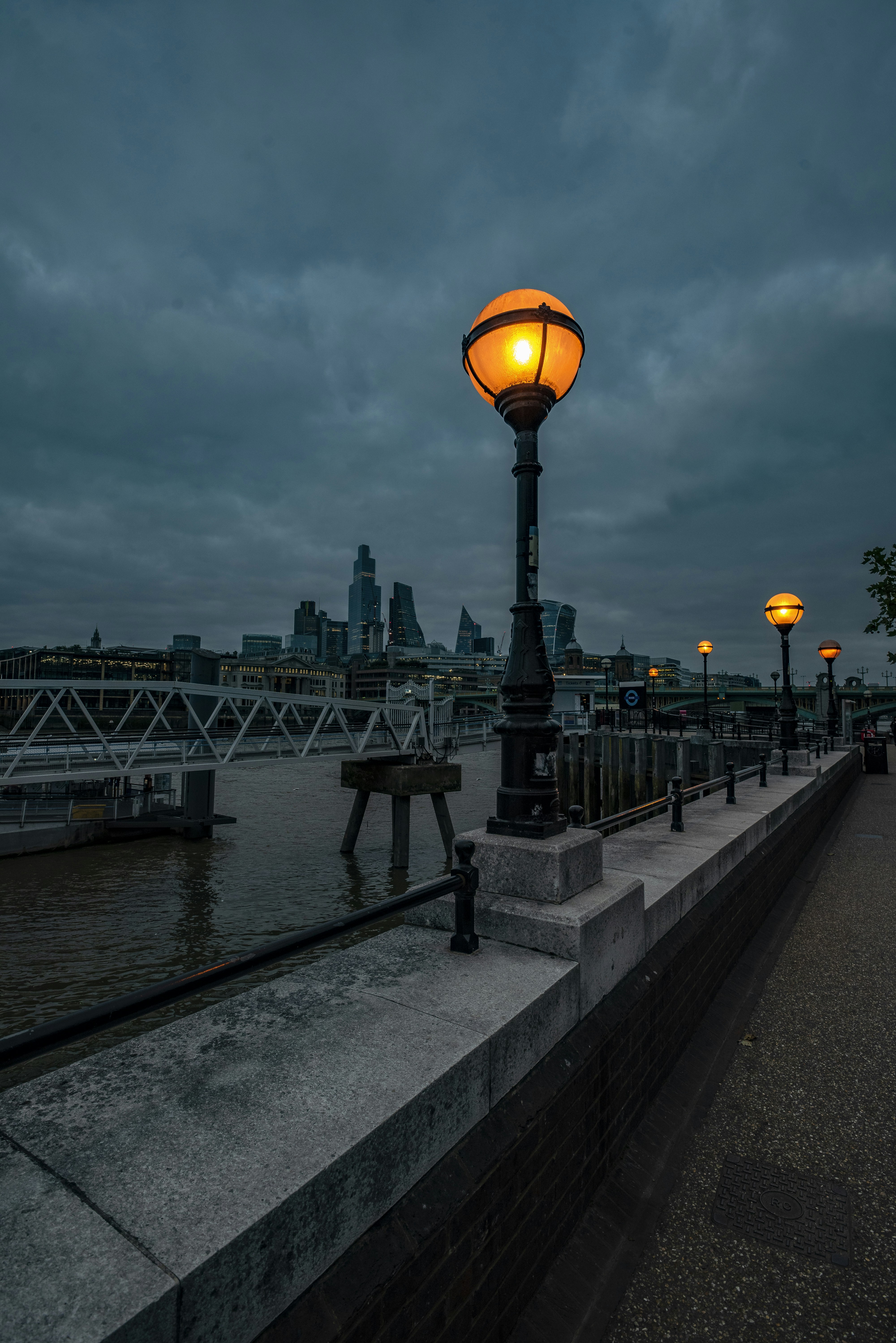 Blue-hour lamps along the riverfront with a distant city skyline
