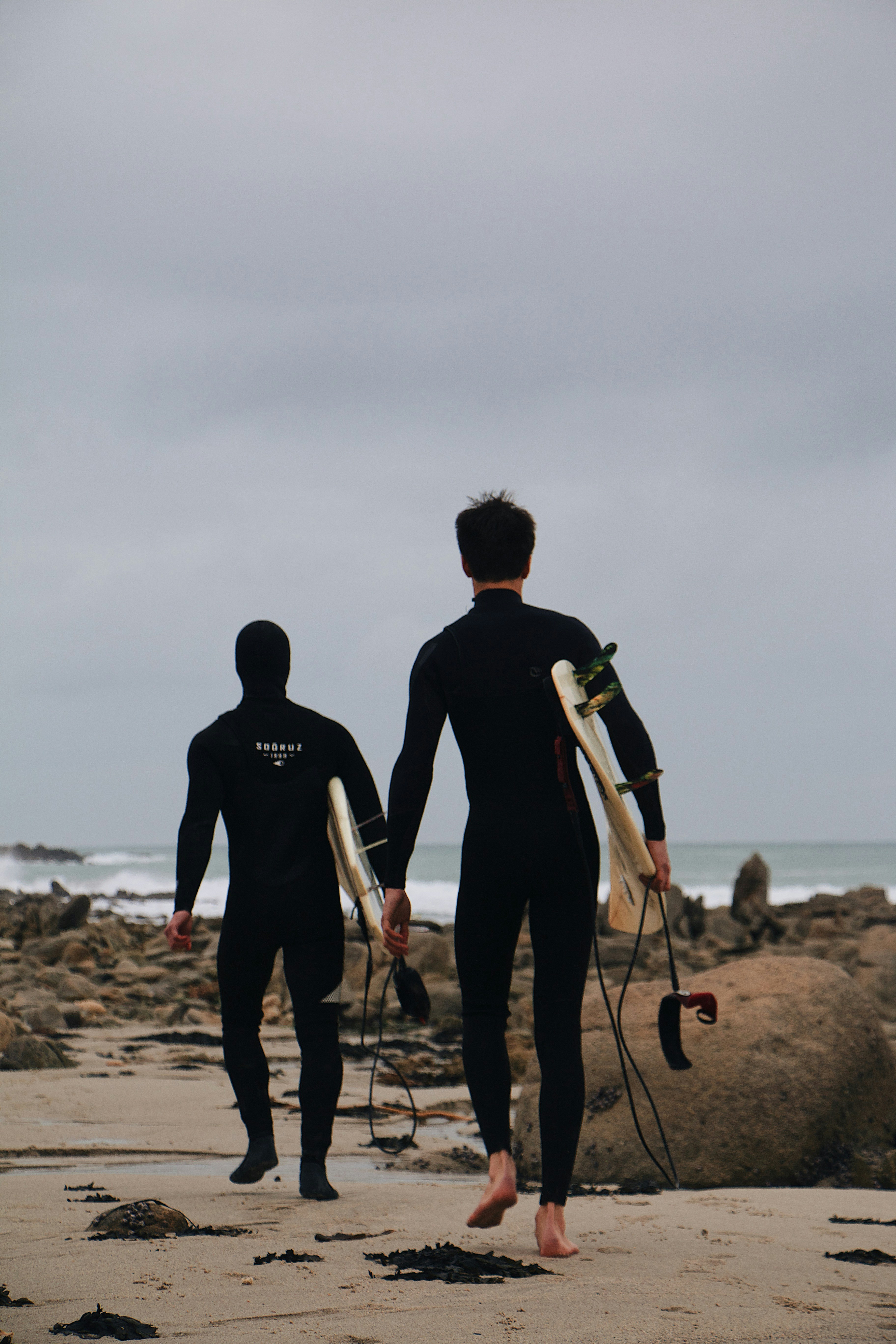 Two surfers in wetsuits walking along a rocky beach, preparing for their next adventure in the ocean. The cloudy sky adds a dramatic backdrop.