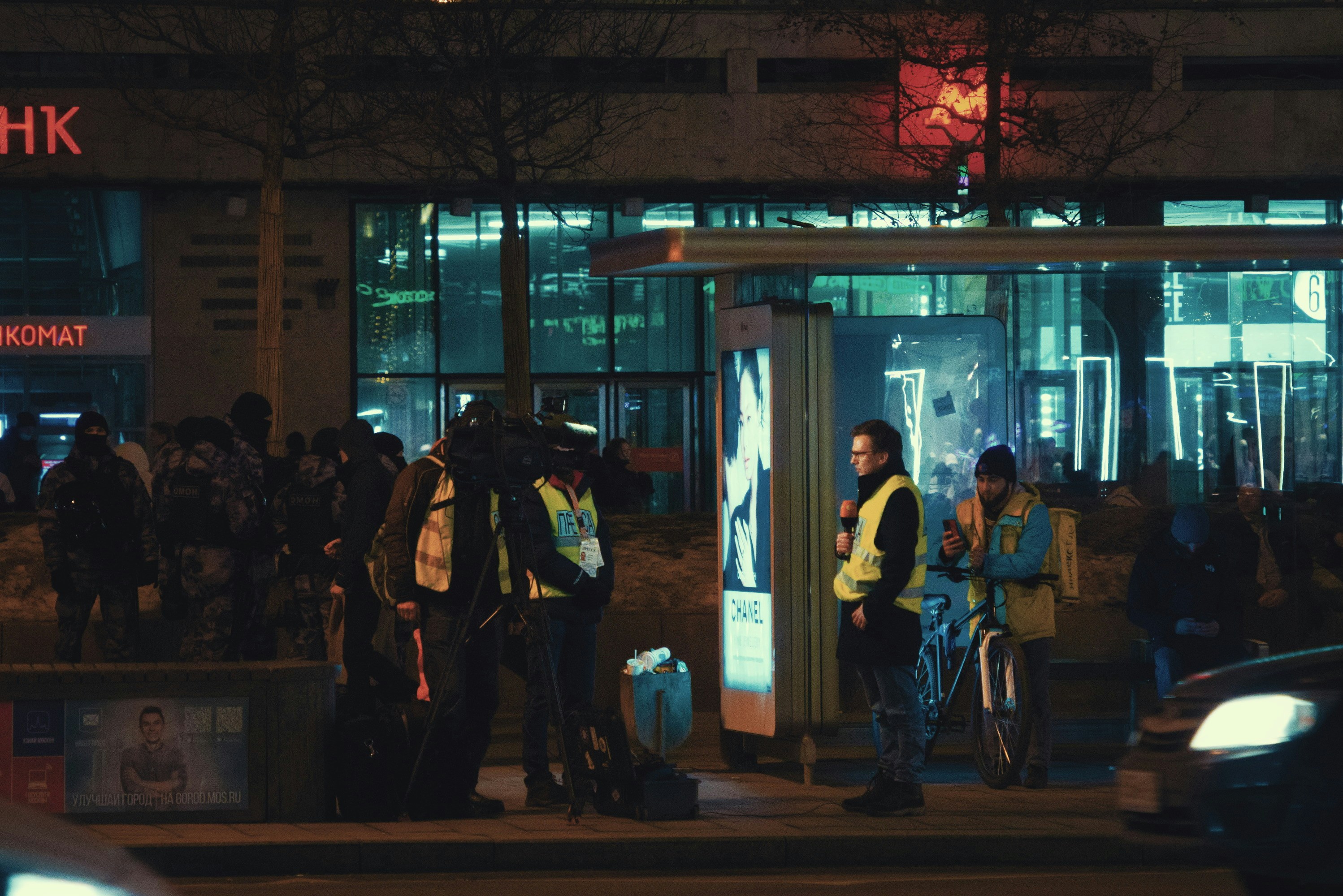 Night-time street vigil: a cluster of people gathered near a bus shelter and a cyclist with a bright vest