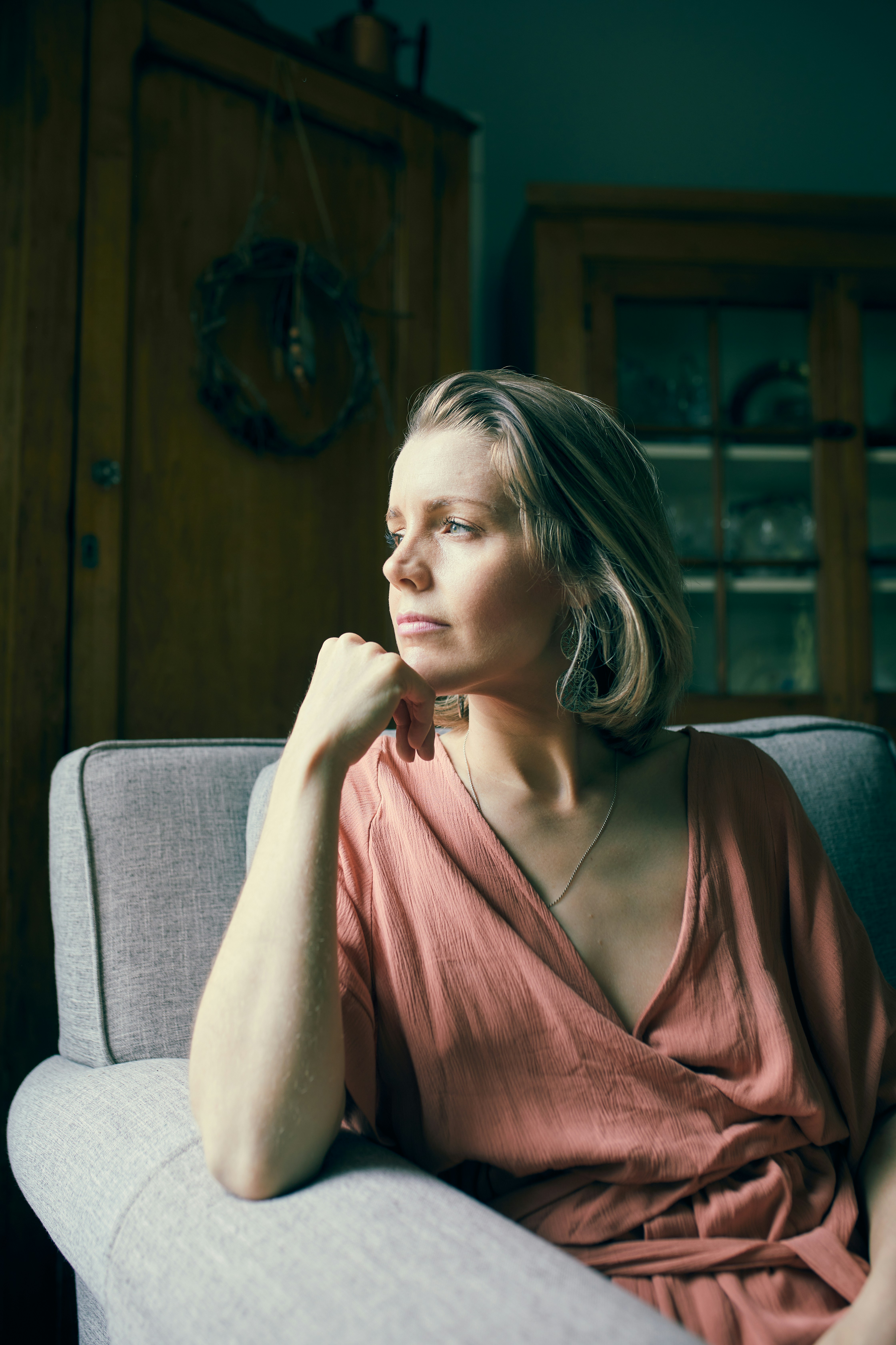 A thoughtful woman gazes into the distance, seated on a gray couch with warm wooden accents in the background.