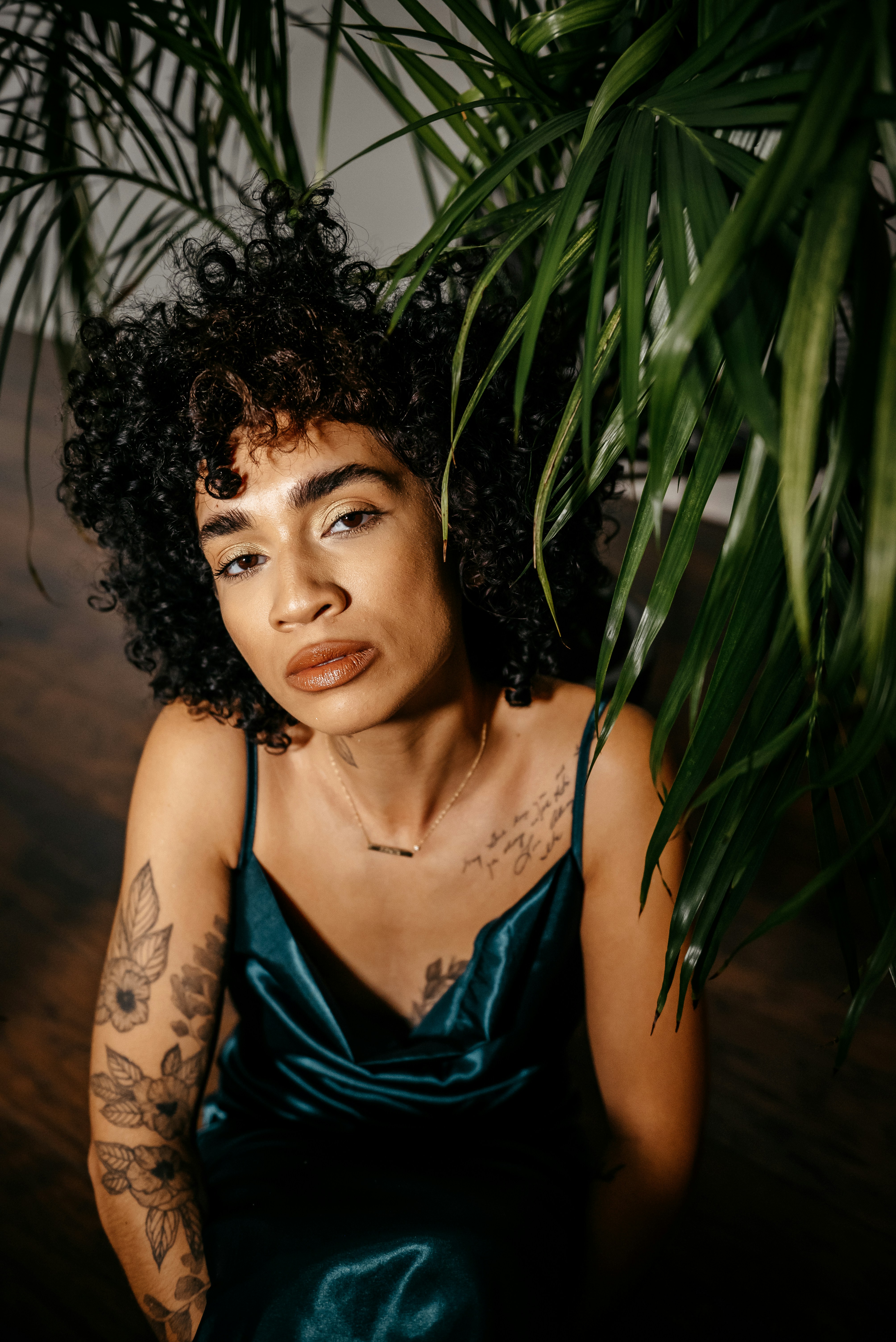 A woman with curly hair and tattoos gazes thoughtfully while surrounded by lush green leaves. Her teal dress contrasts beautifully with the earthy tones of the setting.