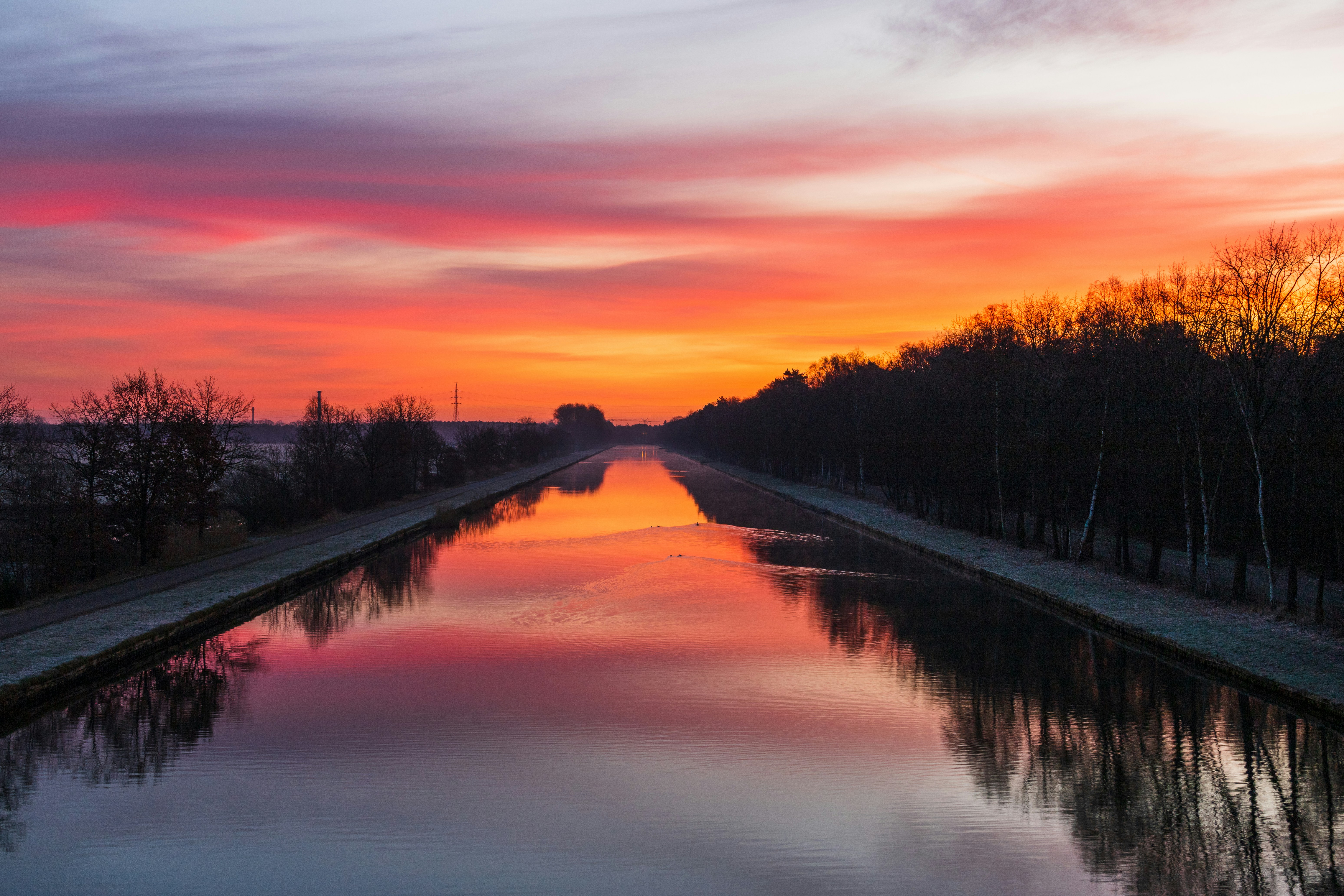 Vibrant sunset reflecting on a tranquil canal, framed by silhouettes of trees along the banks.