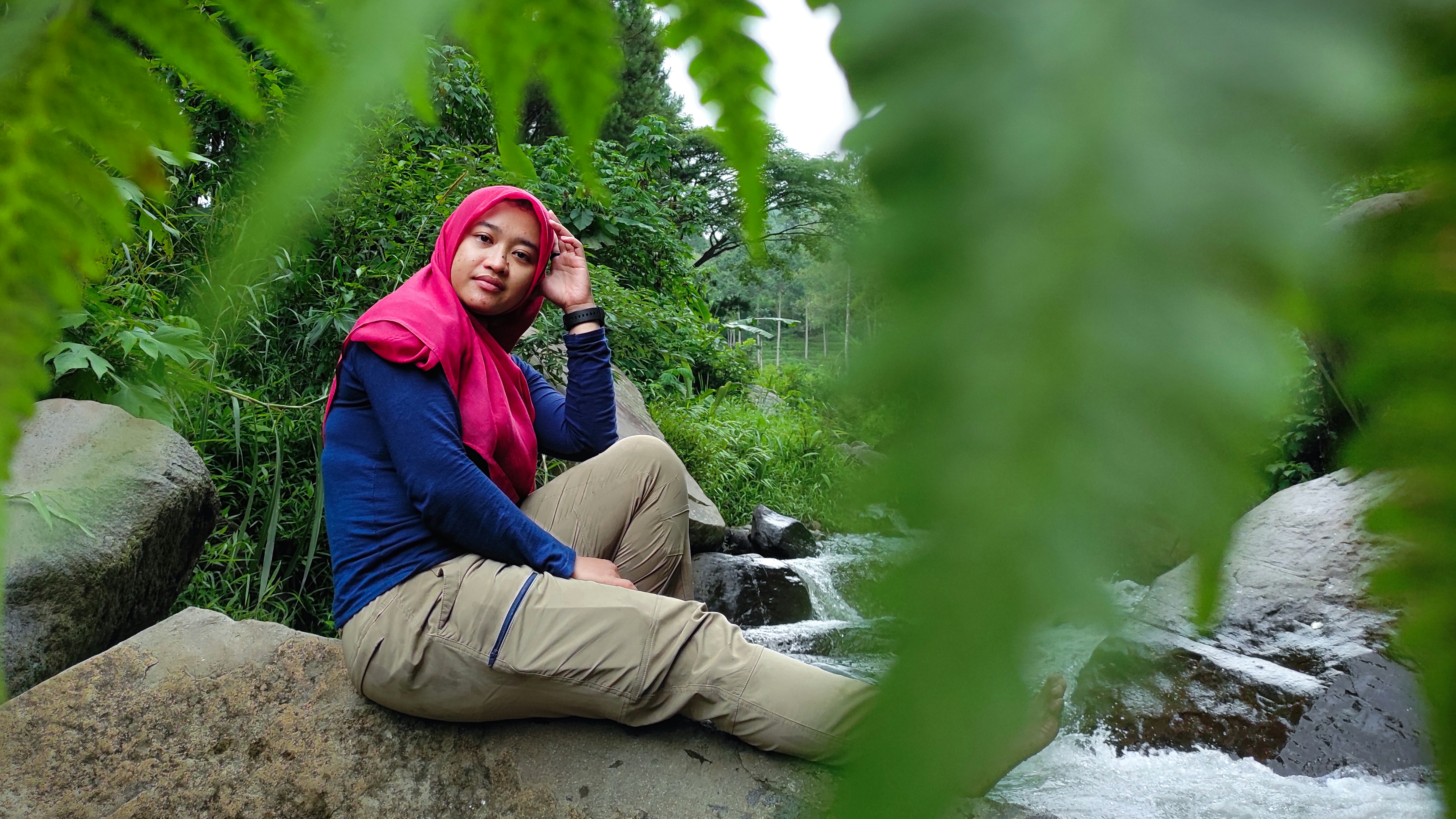 A woman in a pink hijab sits gracefully on a rock by a flowing stream, framed by lush green ferns. The scene captures a moment of tranquility in a vibrant natural setting.