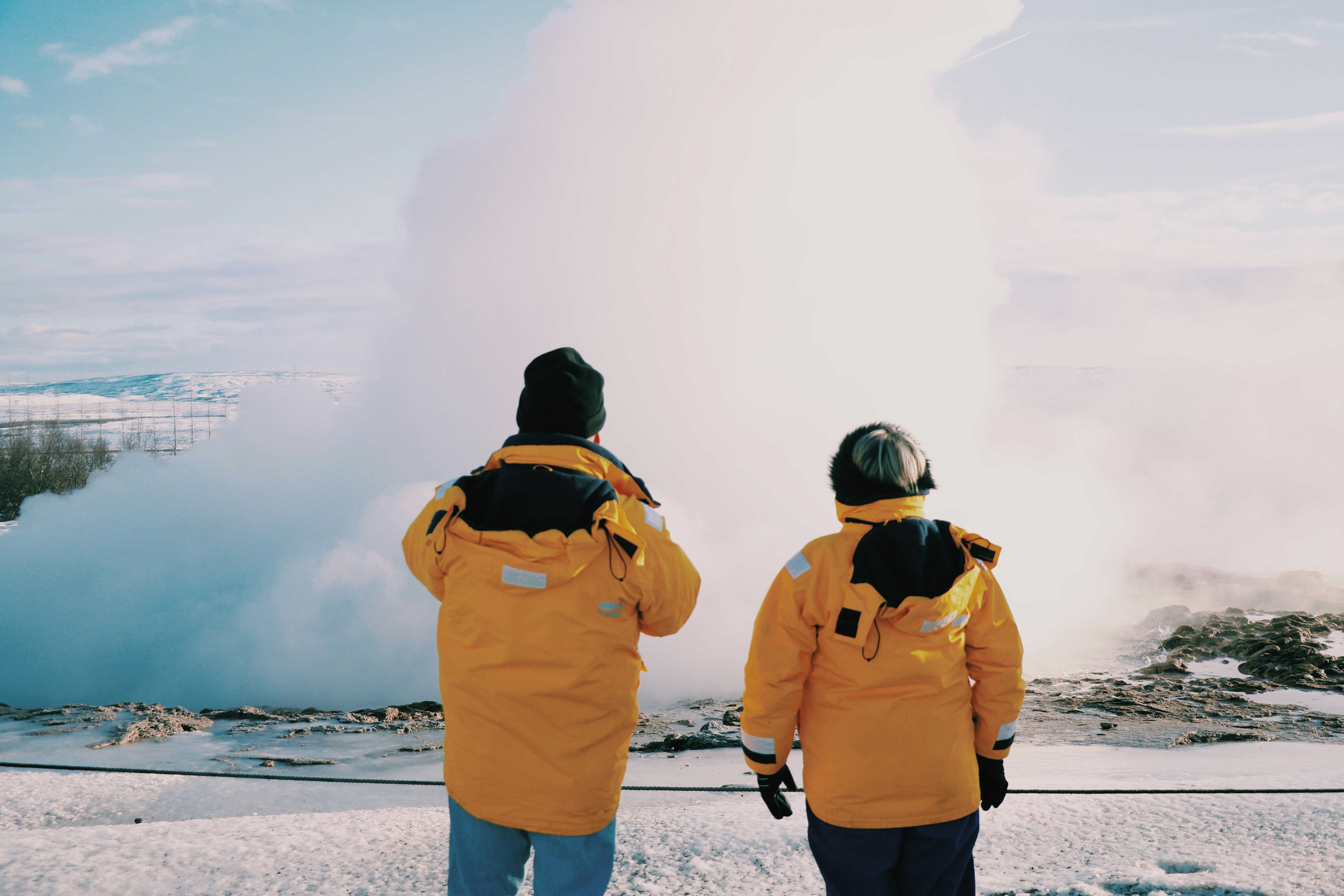Two people in yellow jackets stand before a steaming geyser under a bright sky.