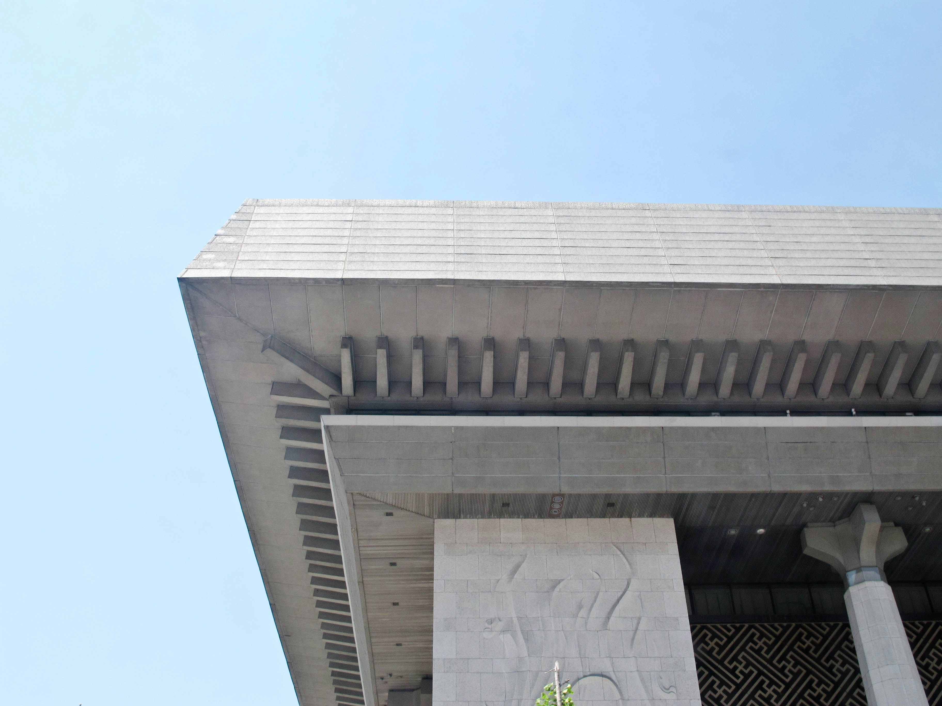 Concrete corner of a modern building with ribbed soffits rises against a clear blue sky.