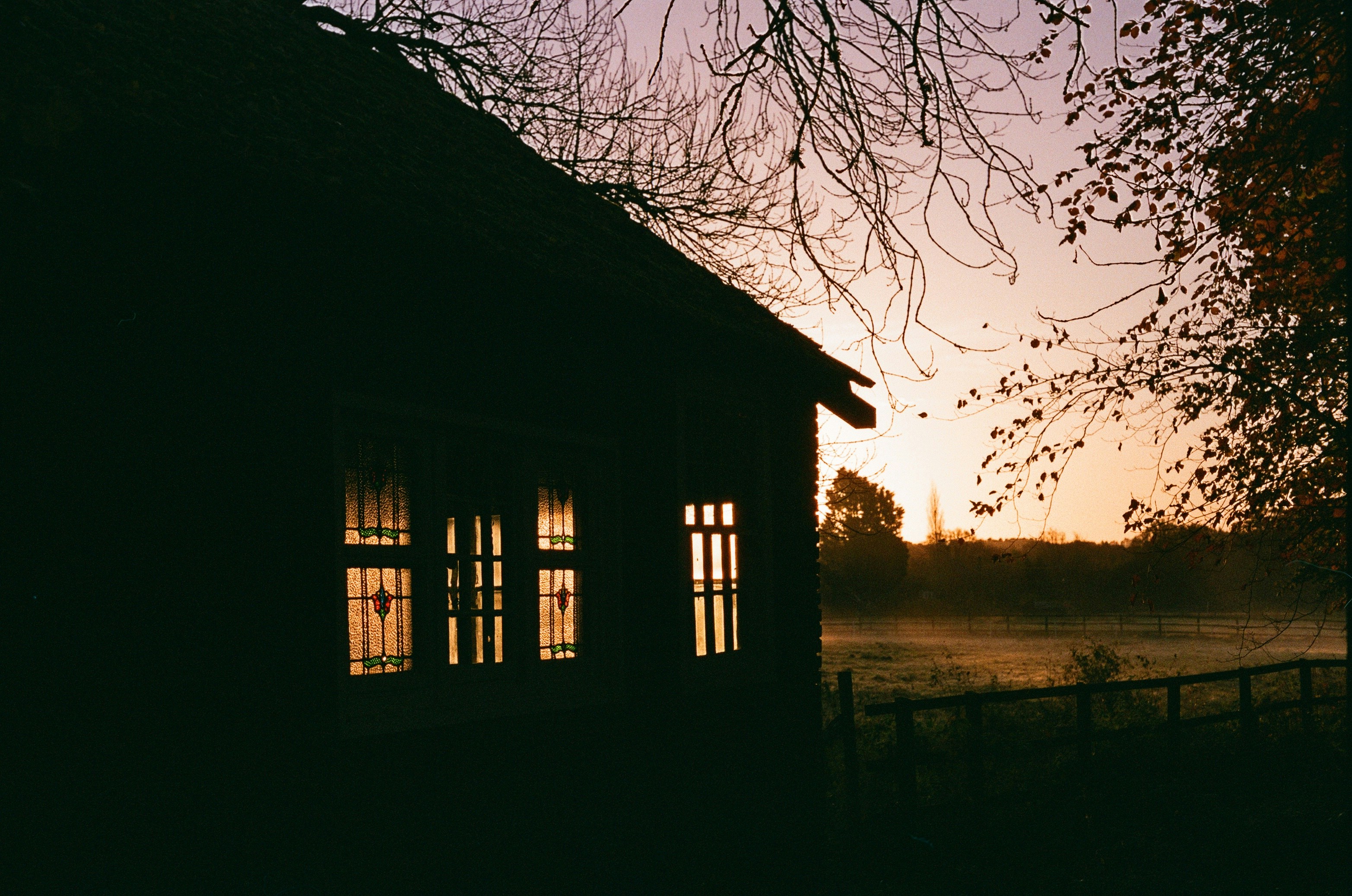 Silhouetted rustic cottage at sunset, warm light glows through the windows as a fence extends into a golden field.