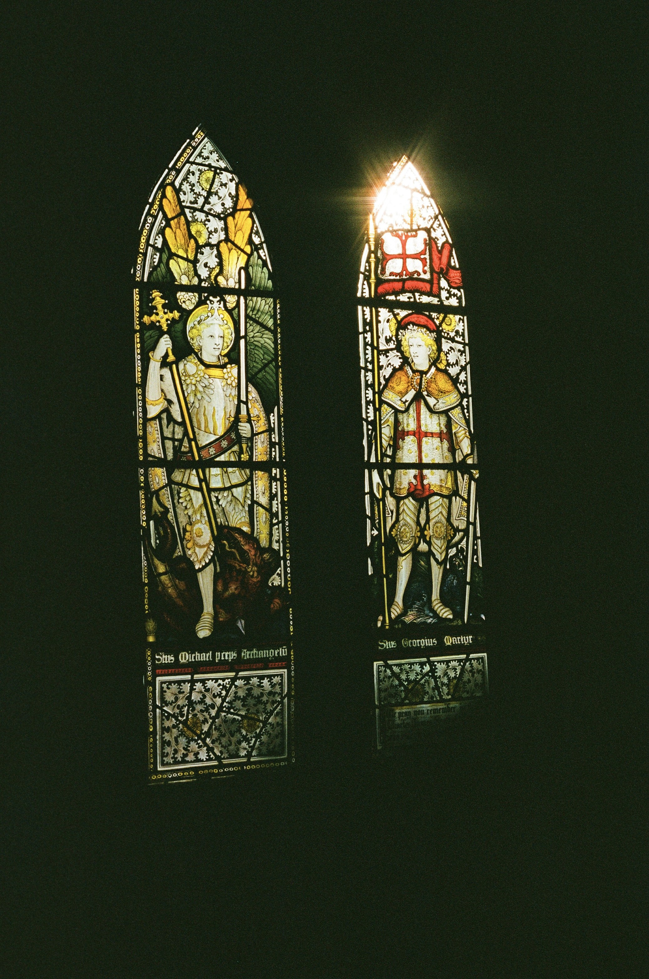 Two stained glass windows depicting historical figures, illuminated by sunlight, set against a dark background.