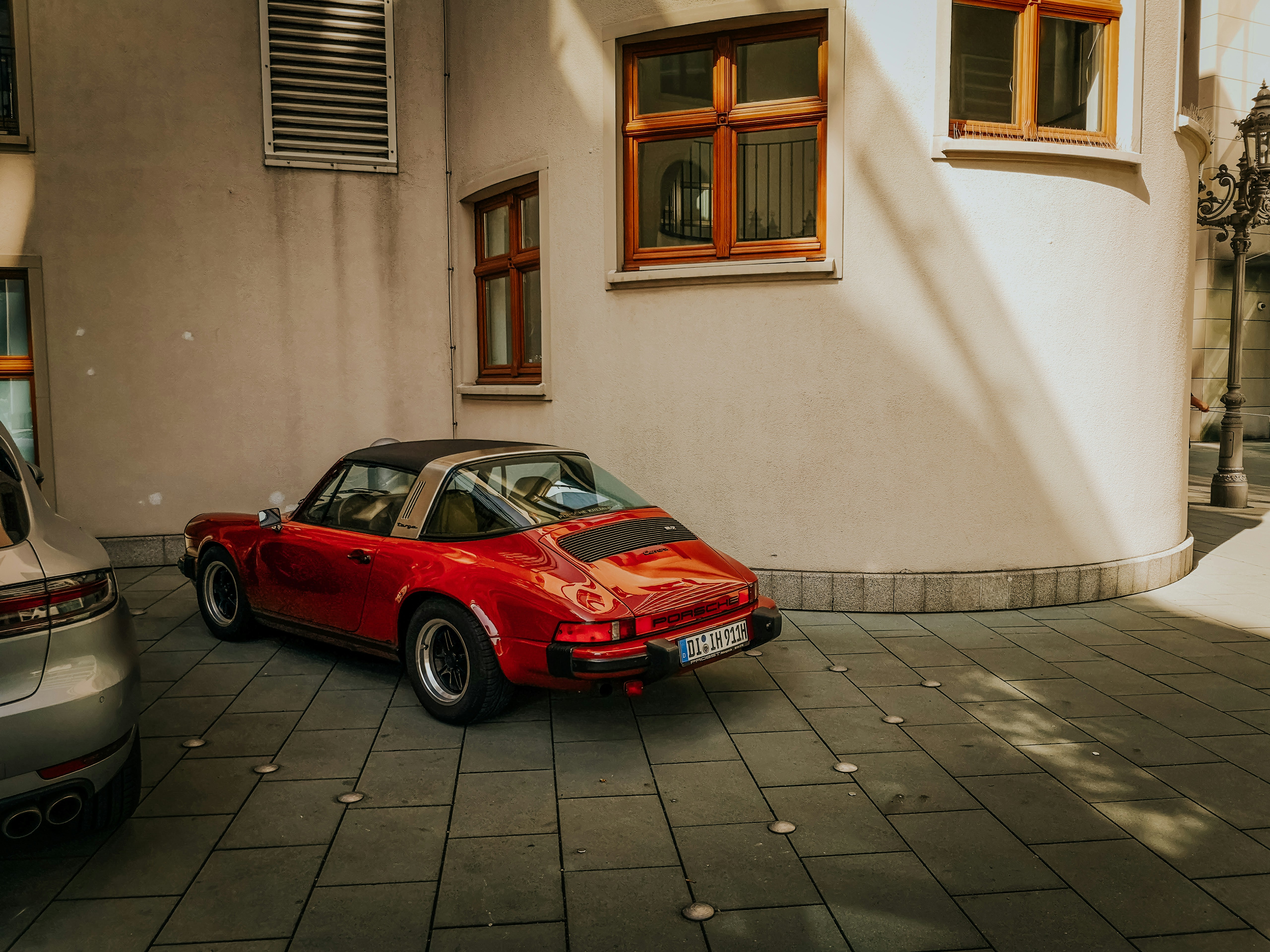 A red vintage Porsche 911 is parked in a sunlit courtyard beside a curved beige wall. The scene emphasizes architectural shadows, tiled paving, and the car's glossy finish.
