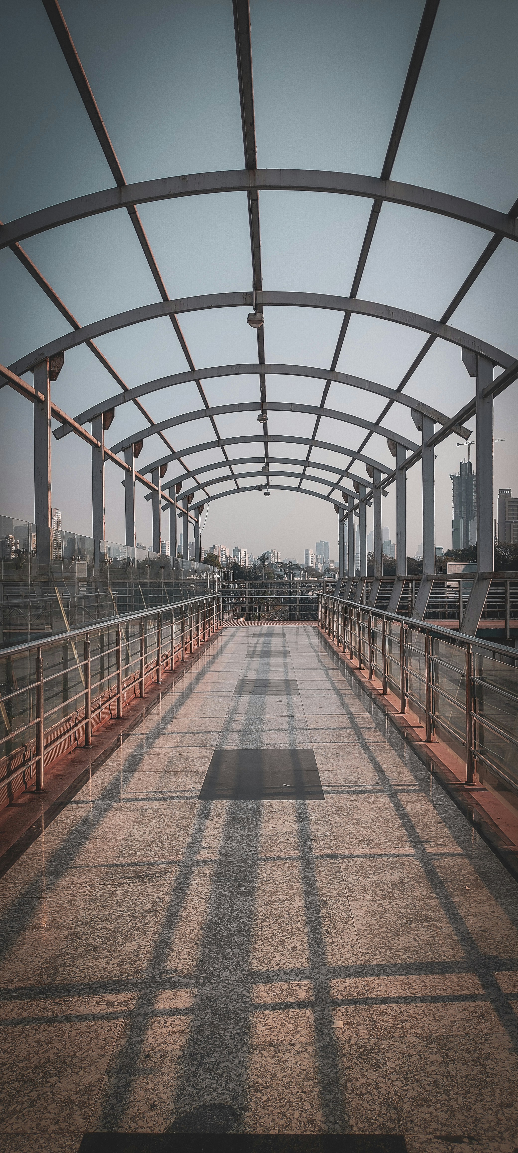 A modern pedestrian walkway framed by a sleek metal canopy, leading towards a city skyline. The interplay of light and shadows adds depth to the scene.