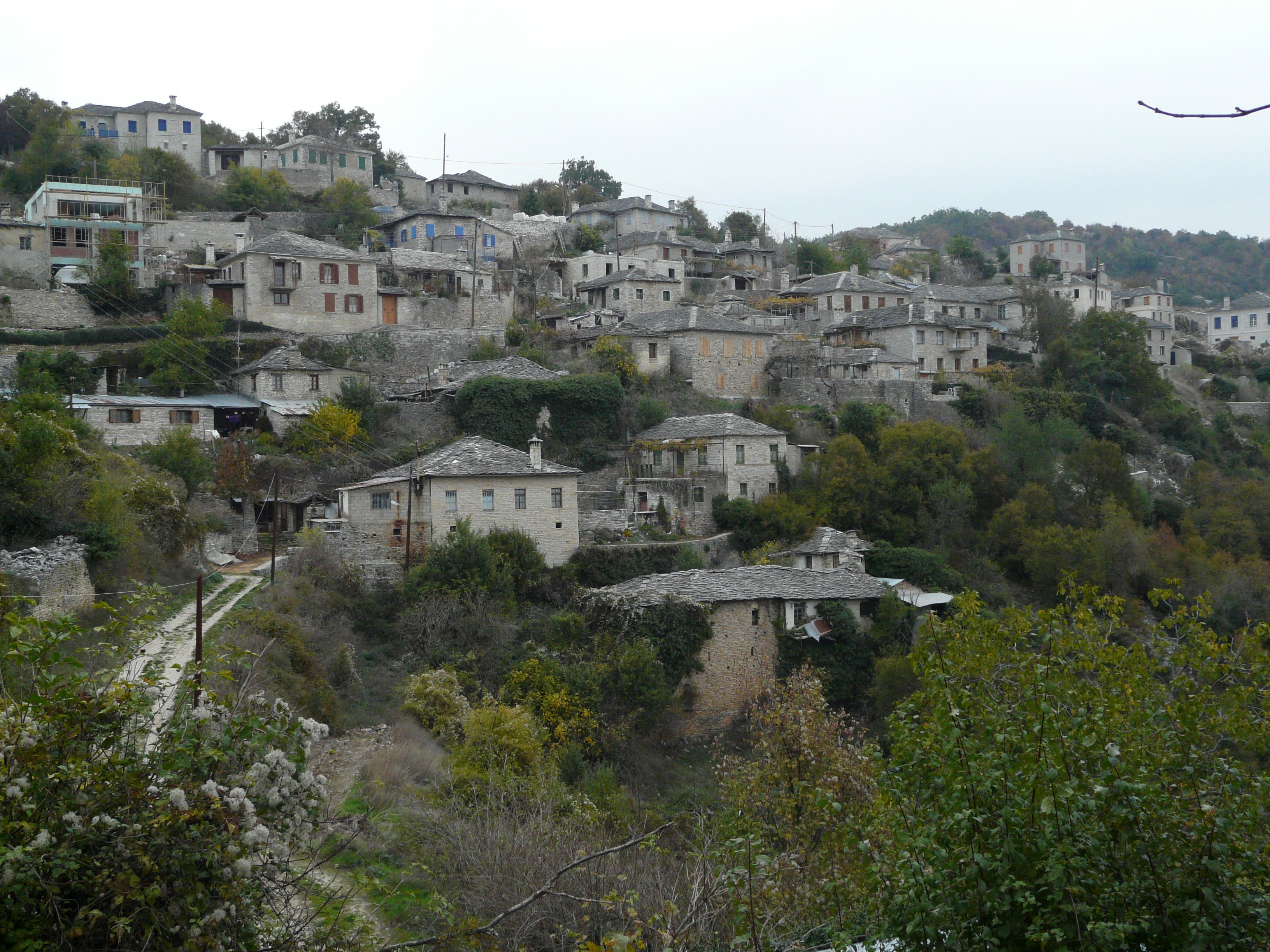 Terraced stone houses cling to a sunken hillside, with olive trees and scrub in the foreground beneath a pale, overcast sky.