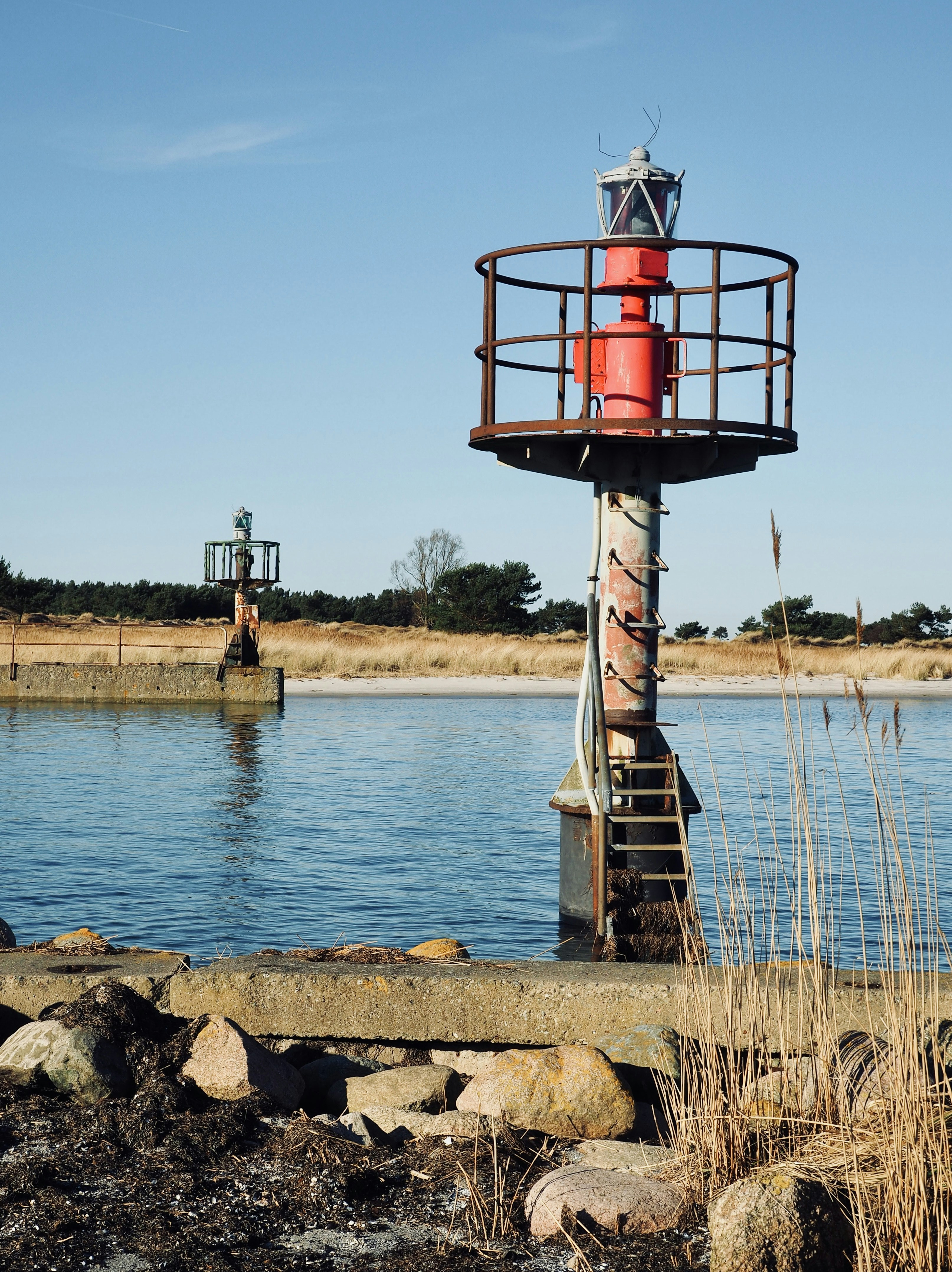 Two lighthouses standing sentinel over calm waters, framed by coastal grasses and a clear blue sky.