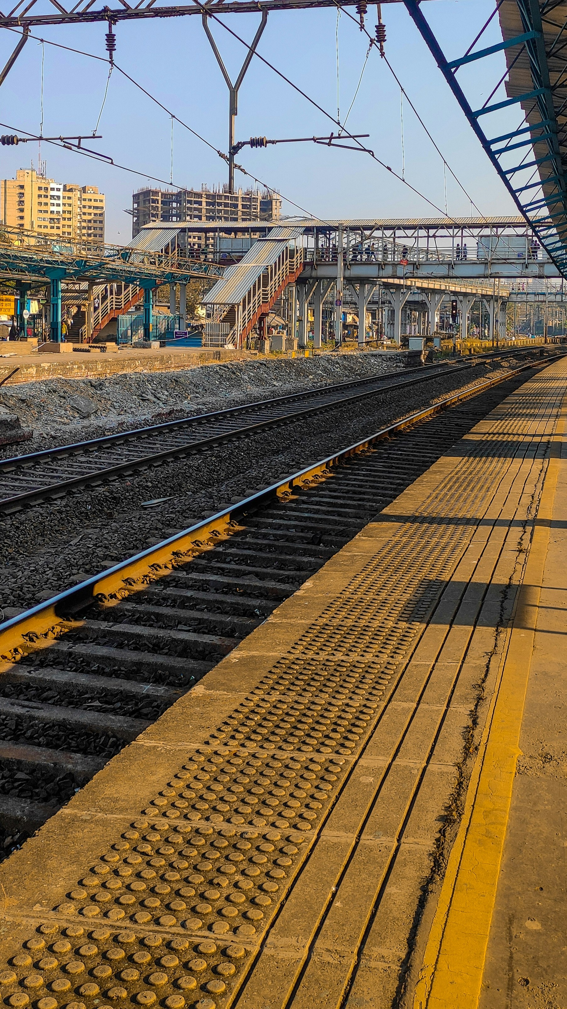 A daytime photograph of a train station platform with yellow tactile paving along the edge, parallel tracks, and a blue steel footbridge overhead.